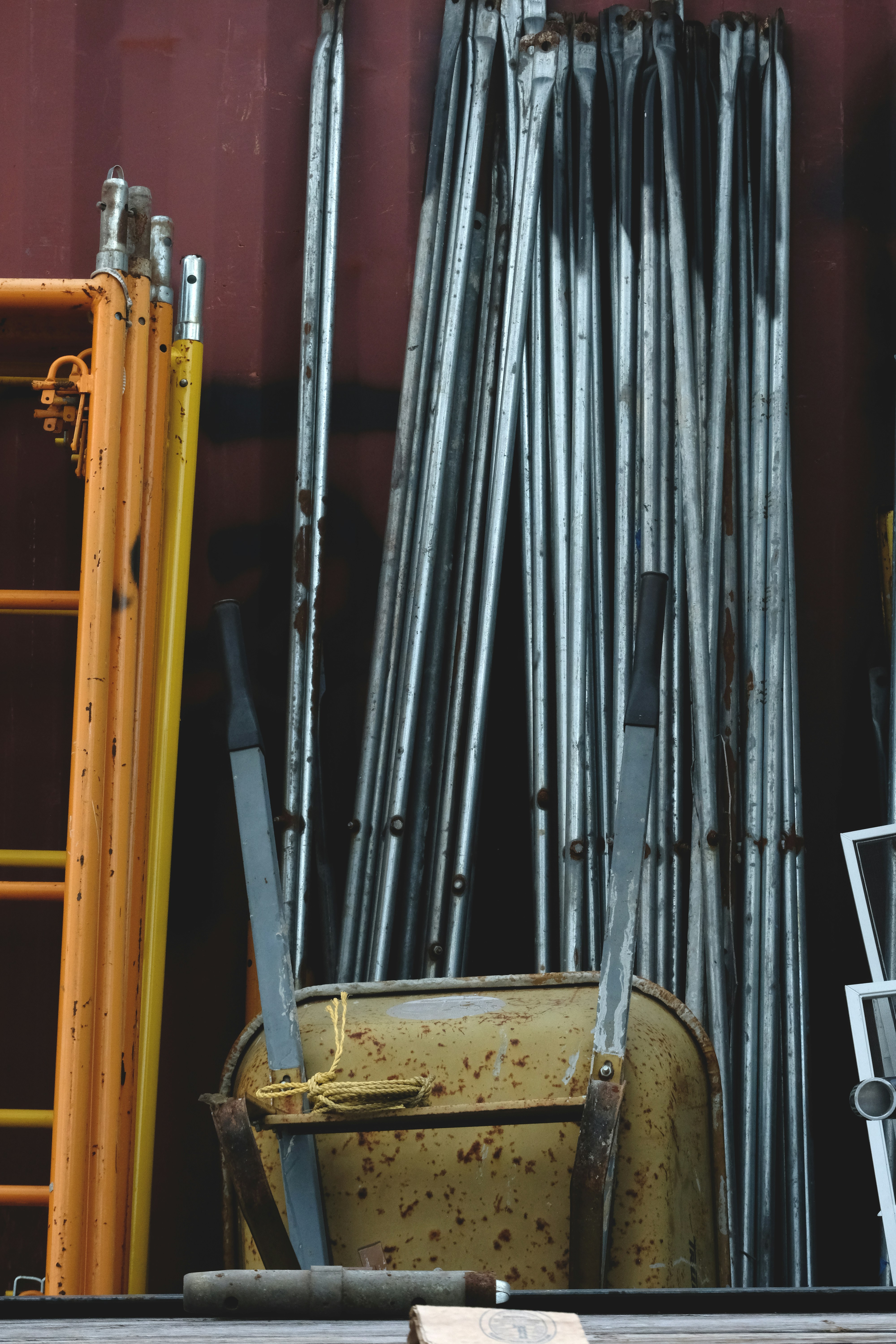 Industrial construction site with orange scaffolding, metal pipes, and yellow wheelbarrow against dark red building wall | Scaffolding and metal poles stacked together