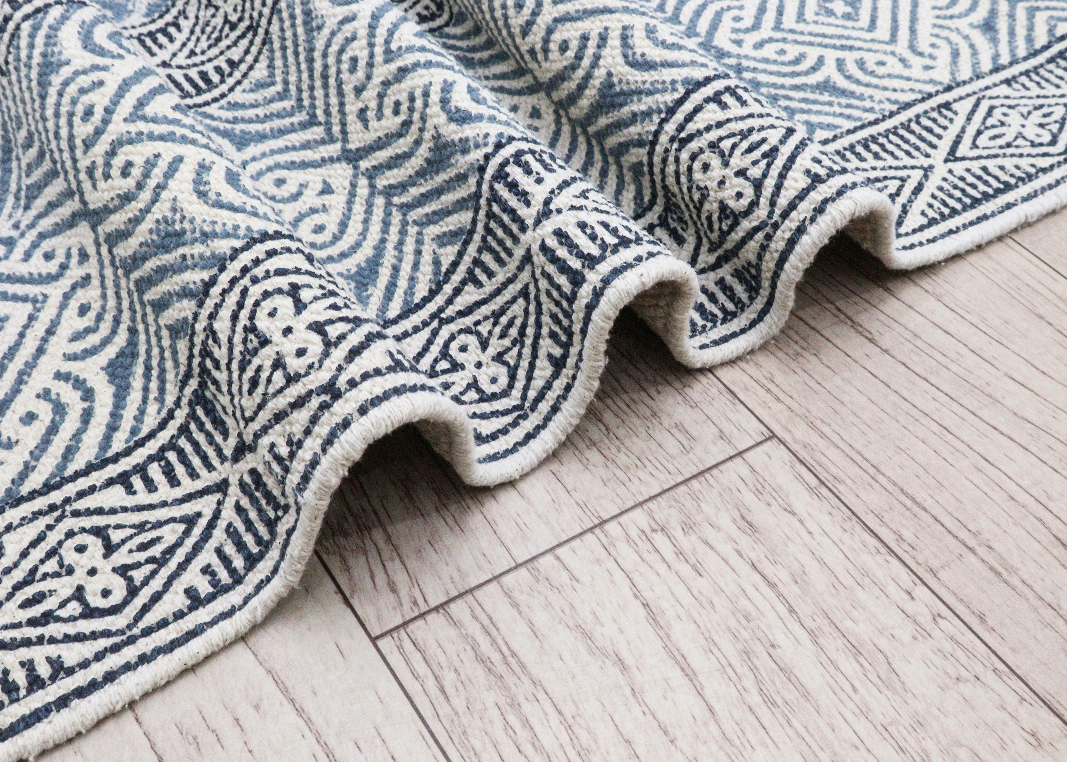 Intricately patterned blue and white rug partially rolled over light wooden flooring, showcasing its texture and design.