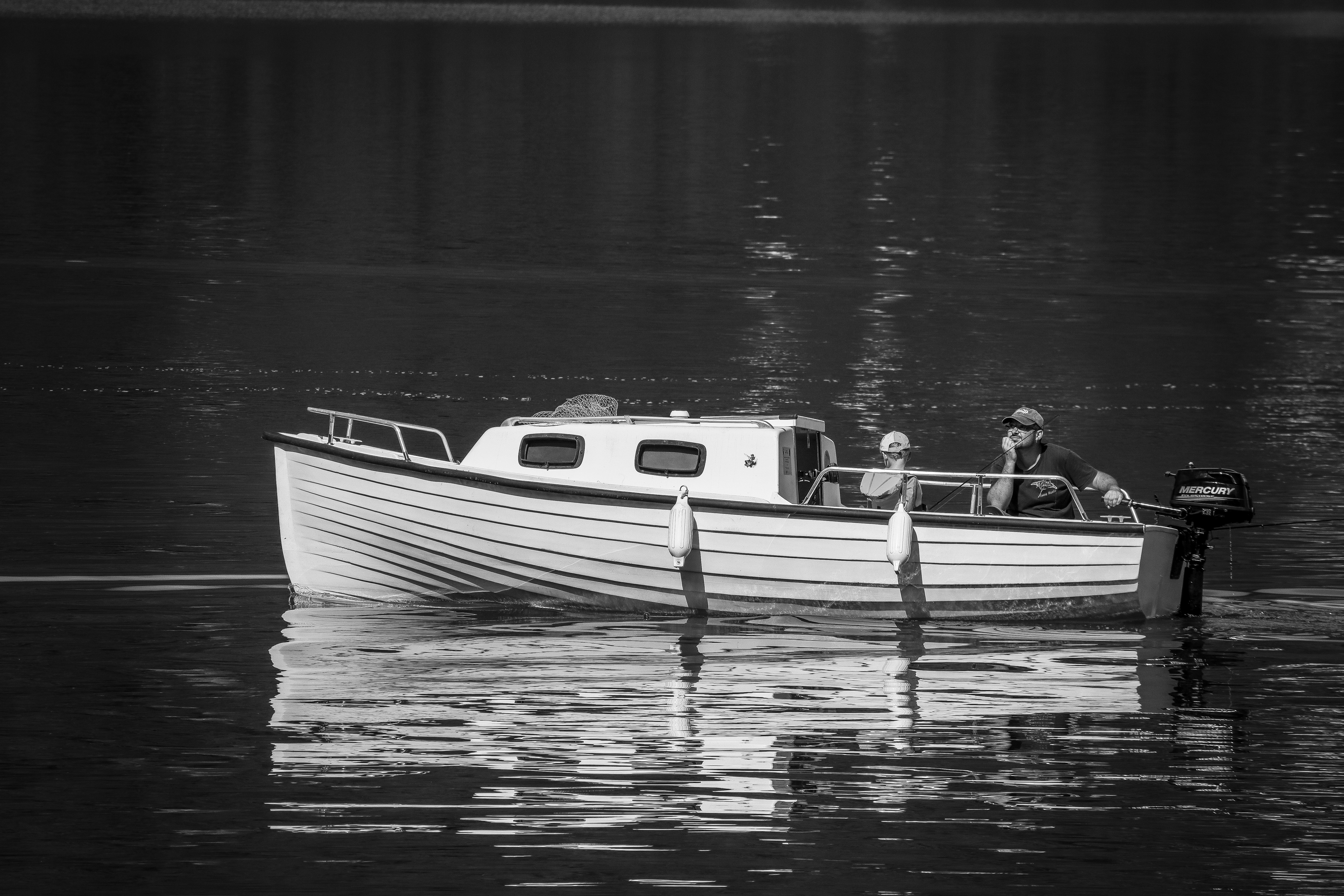 Two individuals in a small boat, casting lines into a calm lake, surrounded by a tranquil atmosphere. Black and white tones enhance the peaceful scene.