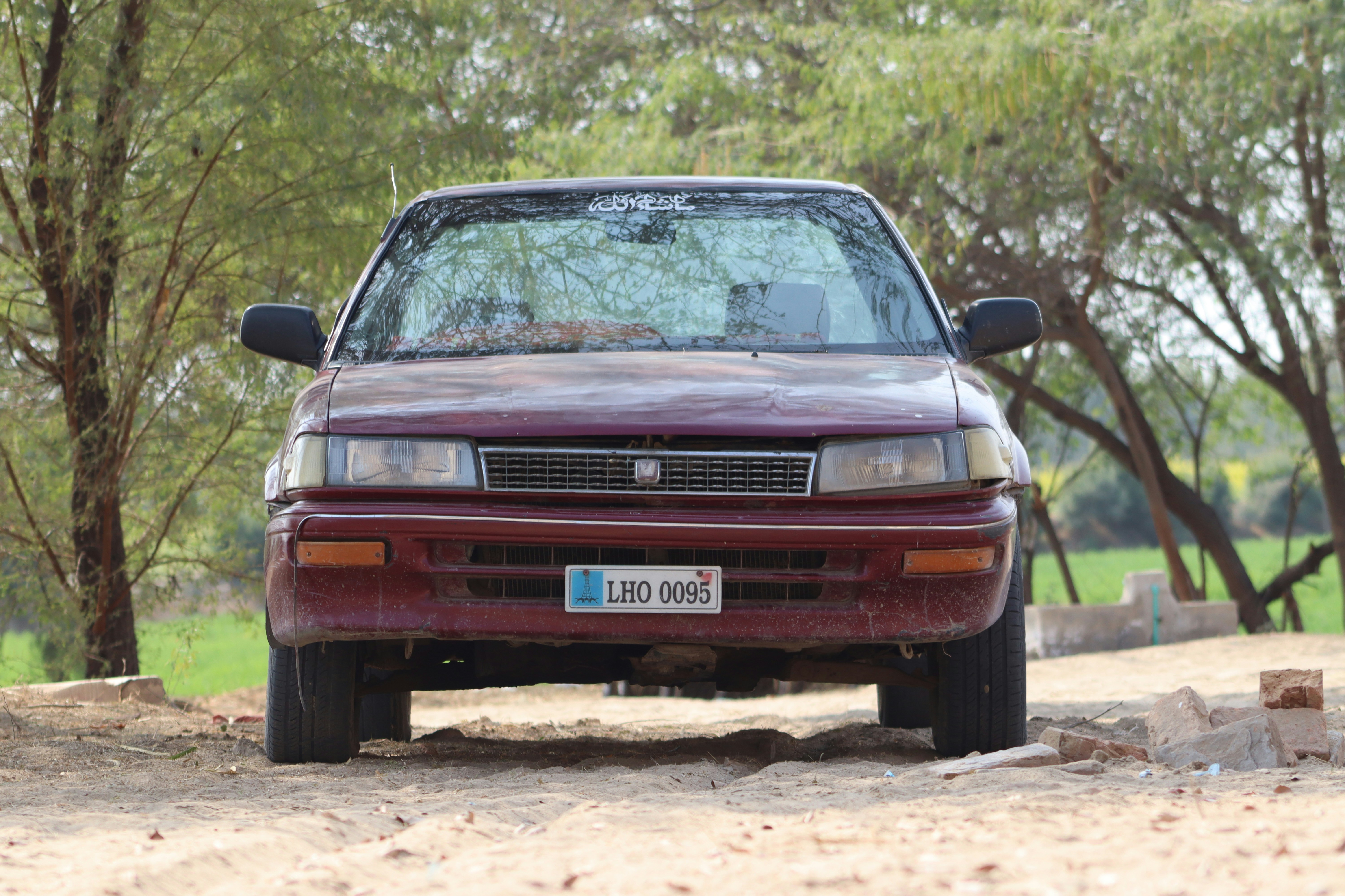 Old maroon sedan parked on a sandy road, framed by lush greenery and distant trees.