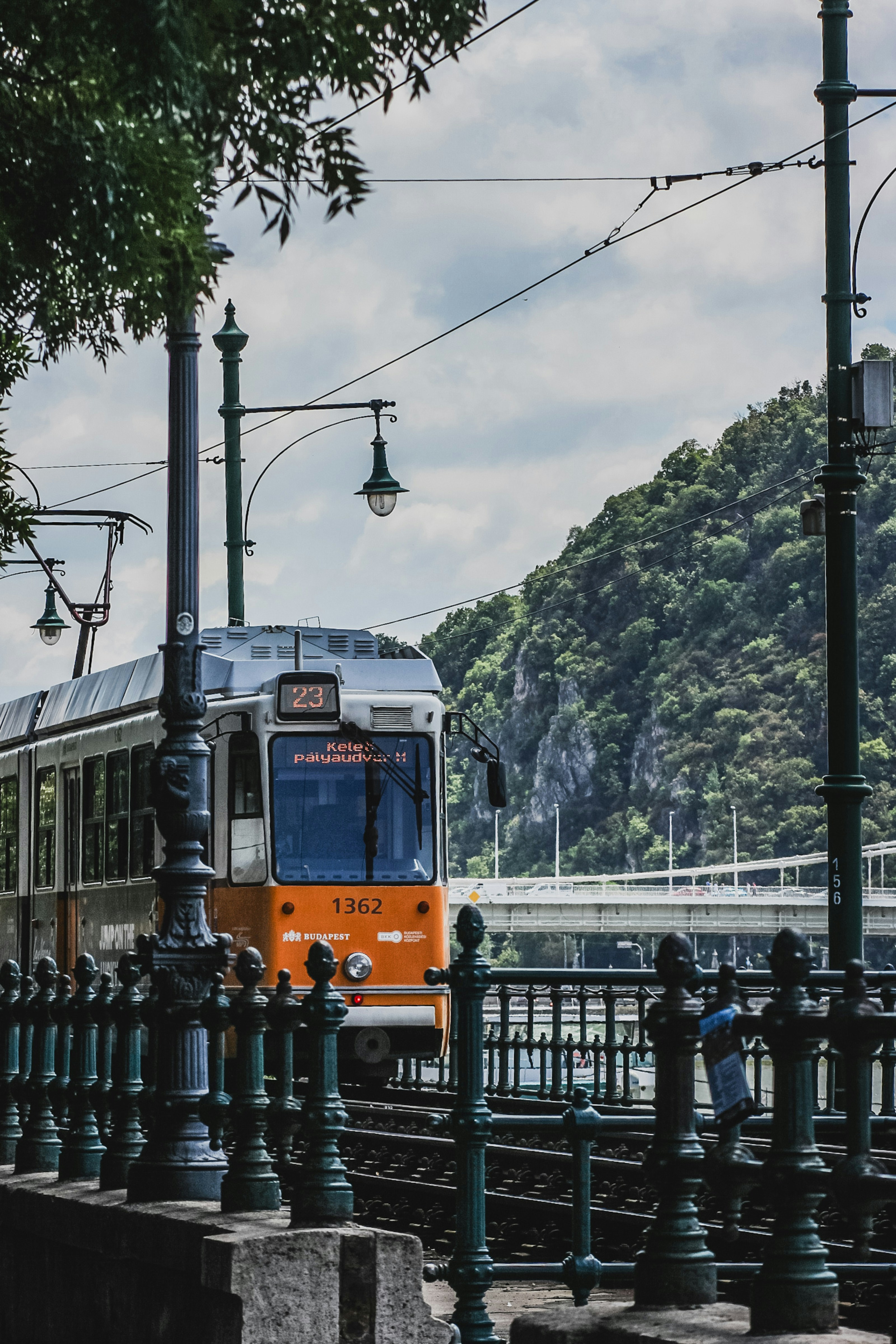 Tram number 23 approaches a stop, framed by ornate streetlamps and lush greenery against a mountainous backdrop.