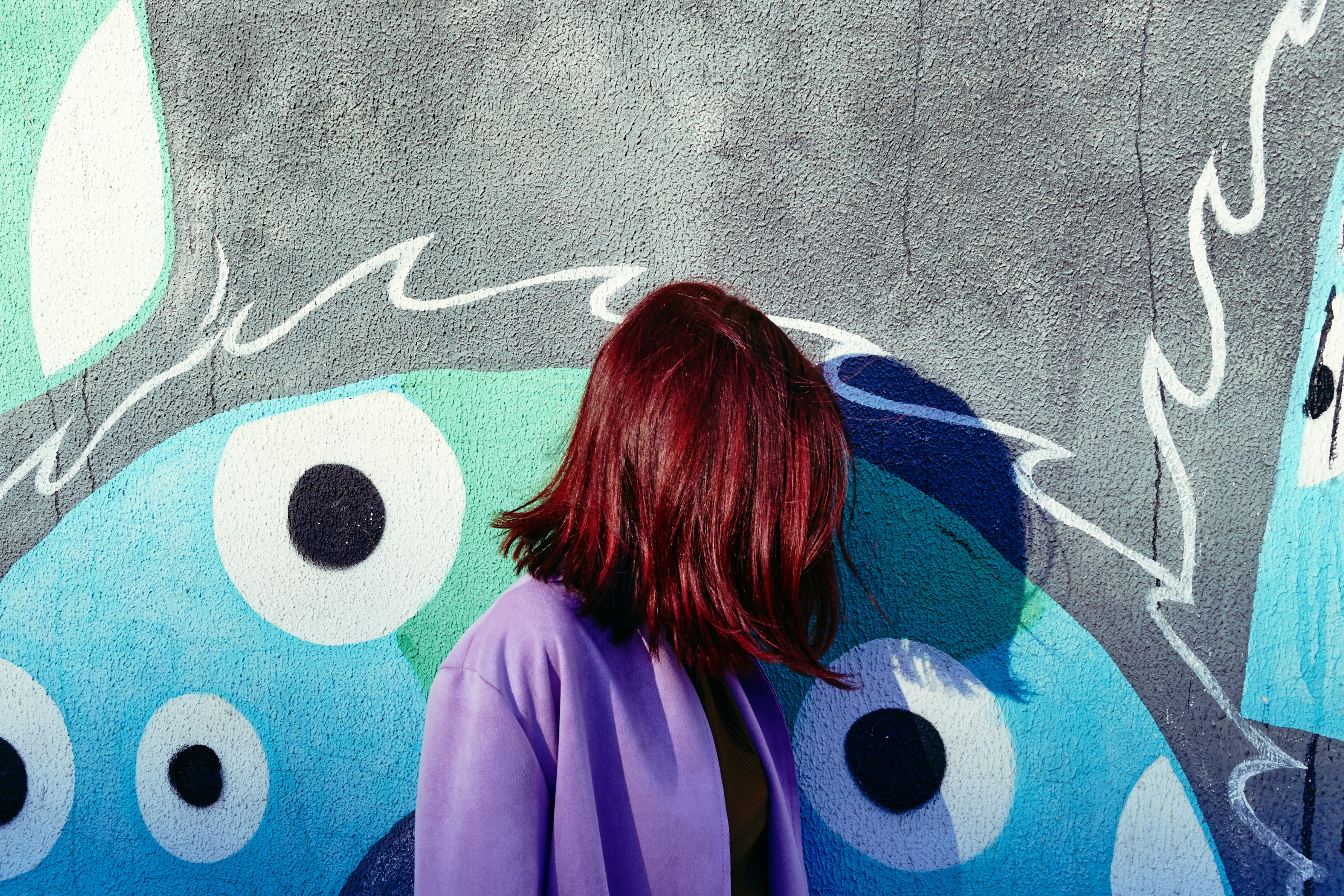 Side profile of a woman with vibrant red hair standing against a painted graffiti wall on a sunny day | Woman with red hair stands before graffiti art