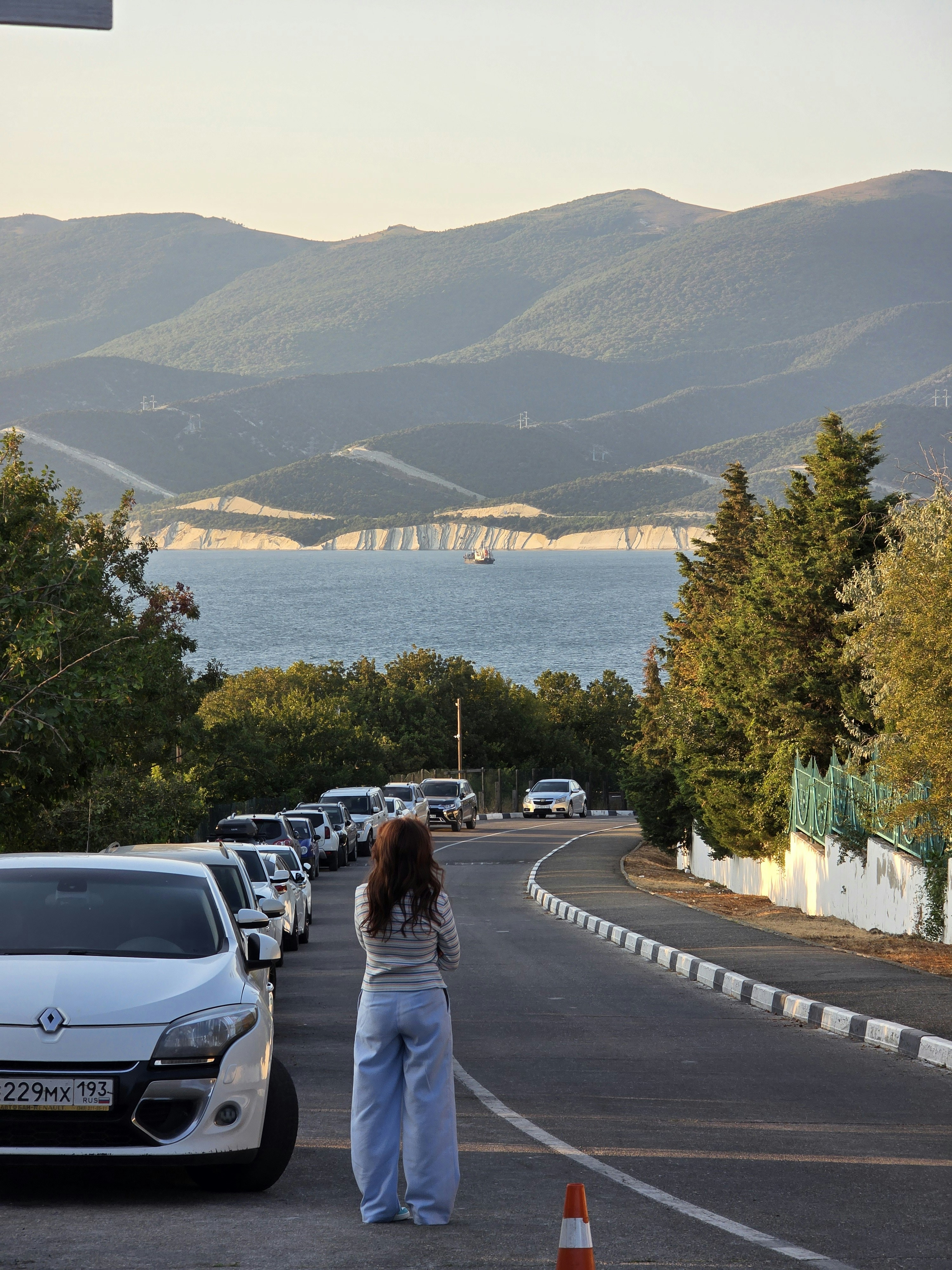A woman stands contemplatively on a winding road, overlooking a tranquil bay framed by rolling hills. The scene captures a moment of peace amidst nature's embrace.