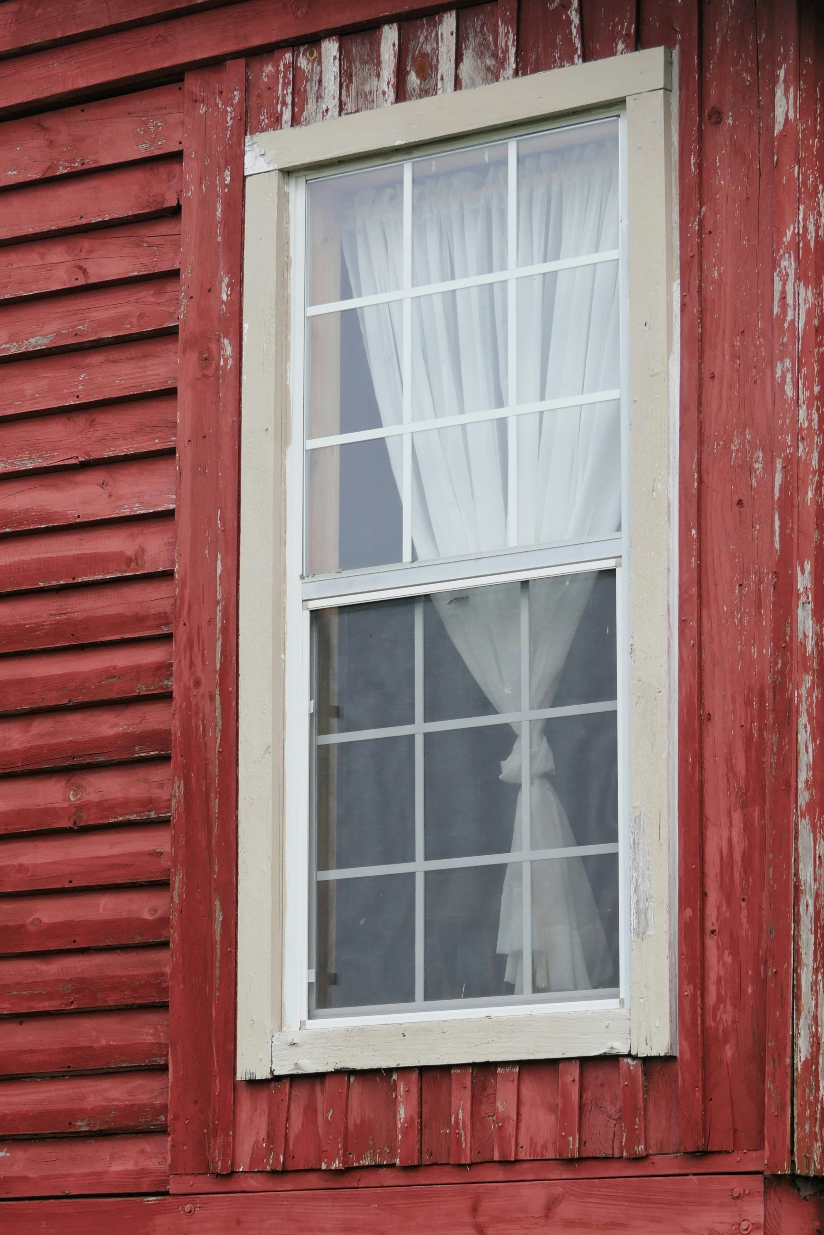 old barn with curtained window | White curtains hang behind a window on a red building.