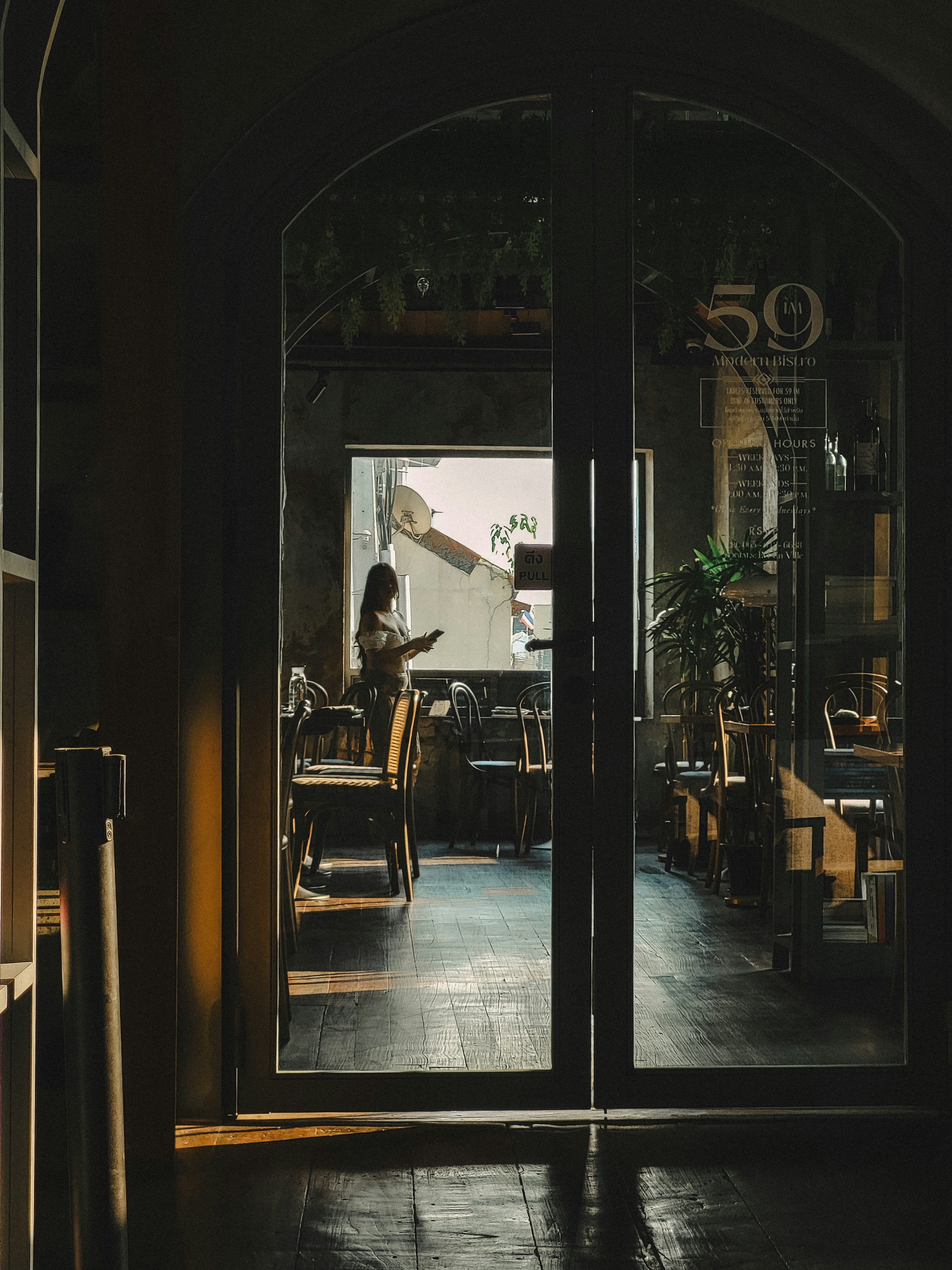 A serene café interior with wooden chairs and a plant, framed by a glass door. A figure stands in the doorway, absorbed in their phone.