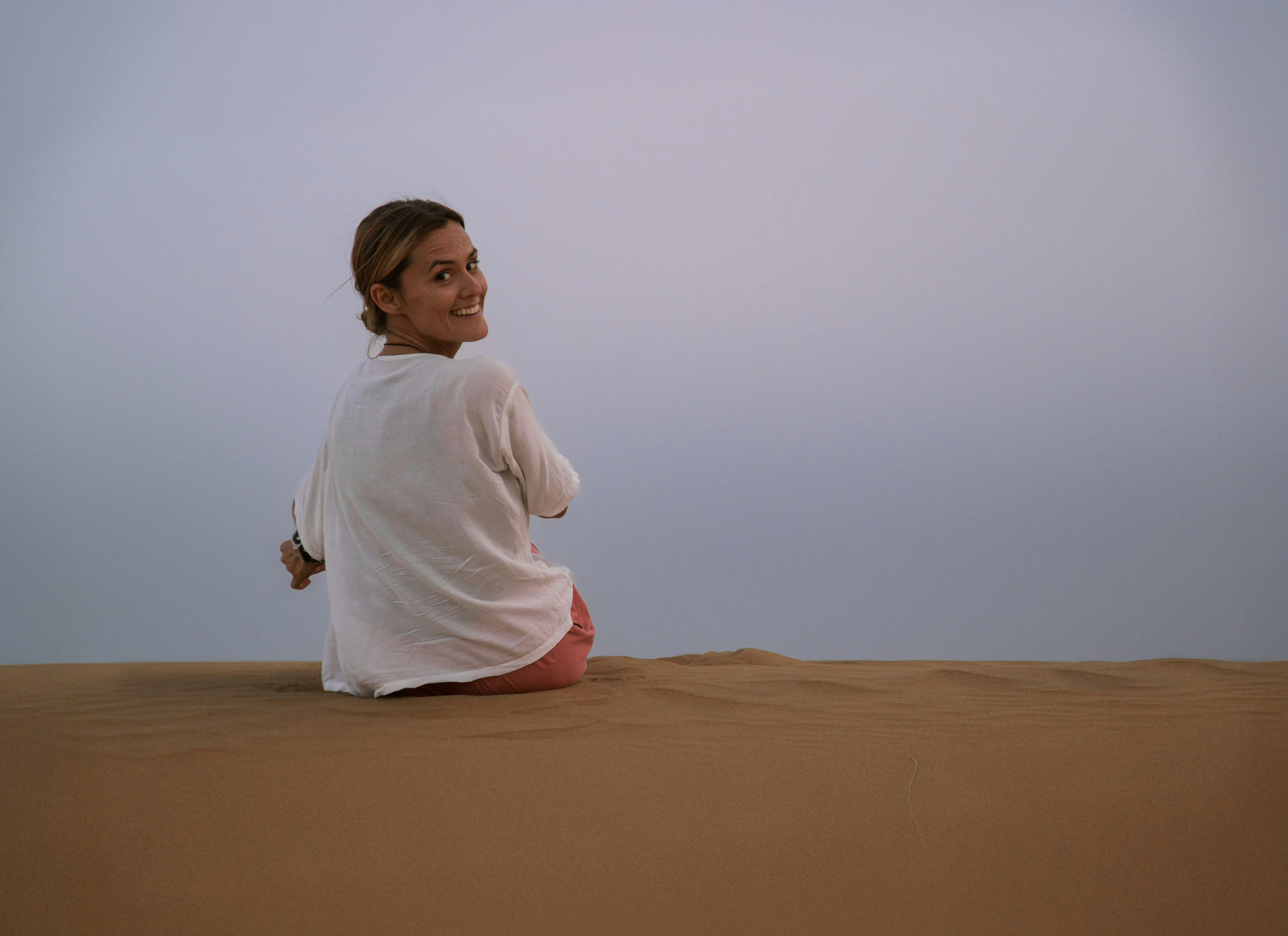 Smiling Woman Sitting on the Dunes at Sunset in Jaisalmer Desert. | Young woman sitting on sand dune looking back