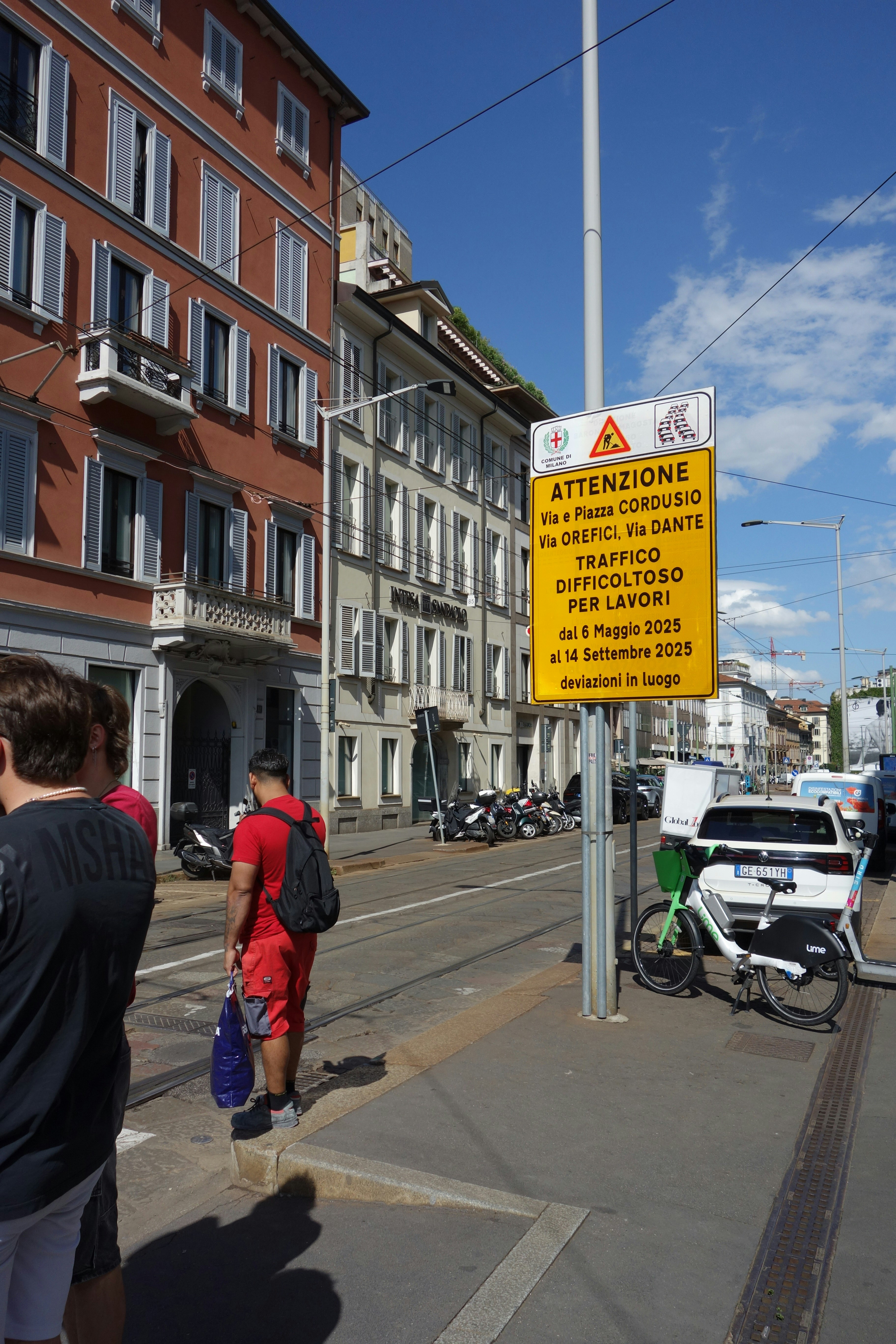 Traffic warning sign indicating construction disruptions on Via Cordusio and Via Orefici, with pedestrians and vehicles in the bustling city scene.