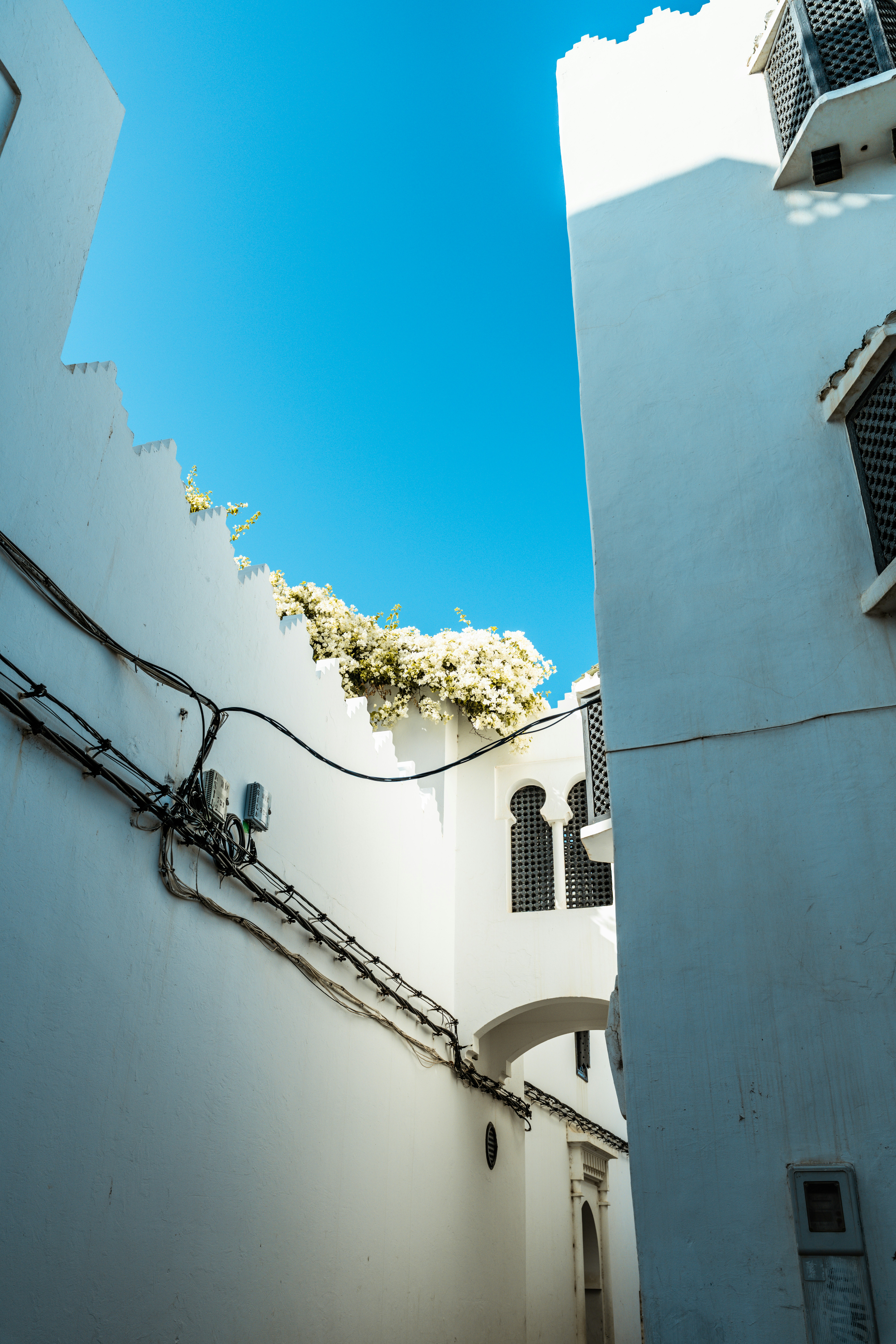 Narrow white-washed alleyway framed by buildings, with vibrant flowers peeking over the edge against a clear blue sky.