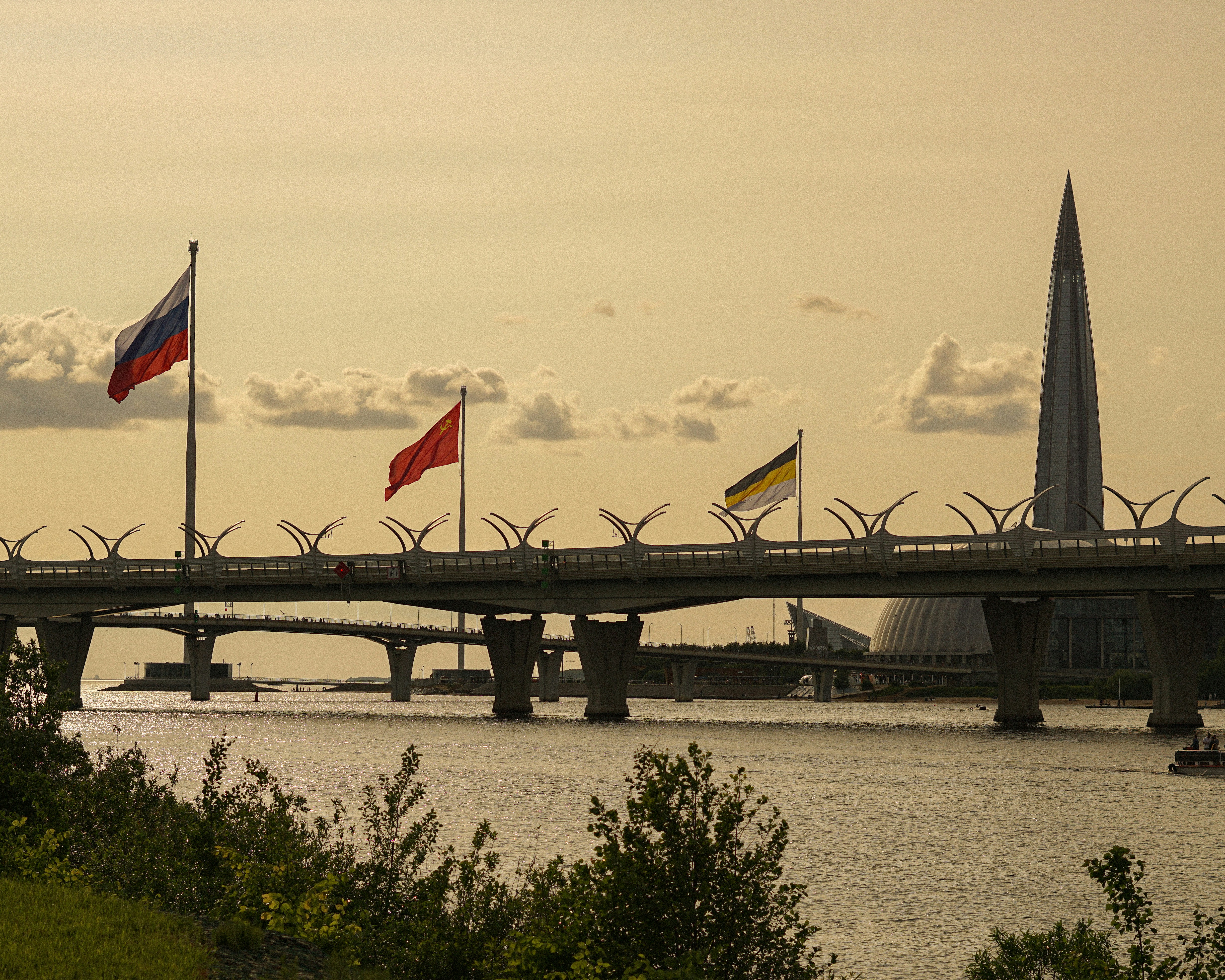 Flags fluttering on a bridge with a modern spire in the background under a golden sky.