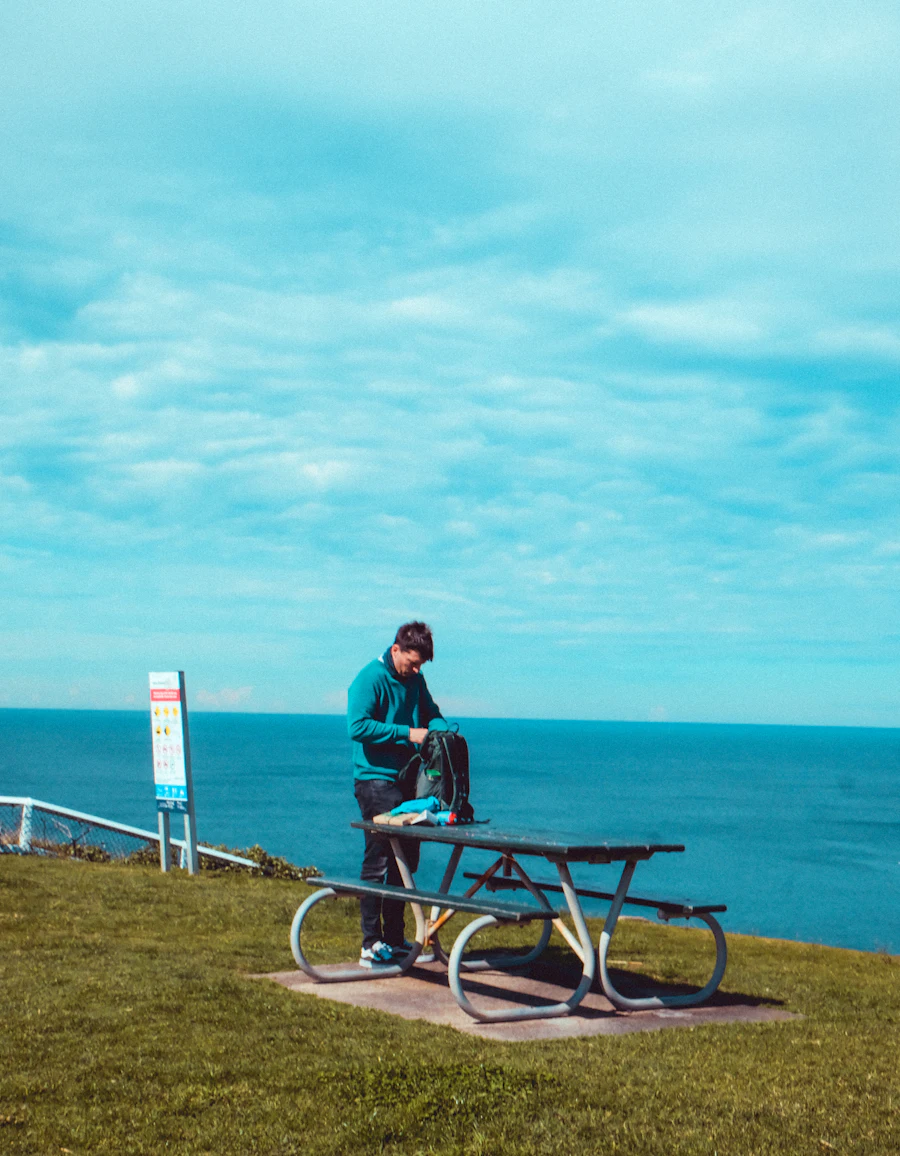 Man packing a backpack at a picnic table on a grassy cliff overlooking a calm blue sea beneath a cloud-streaked sky.