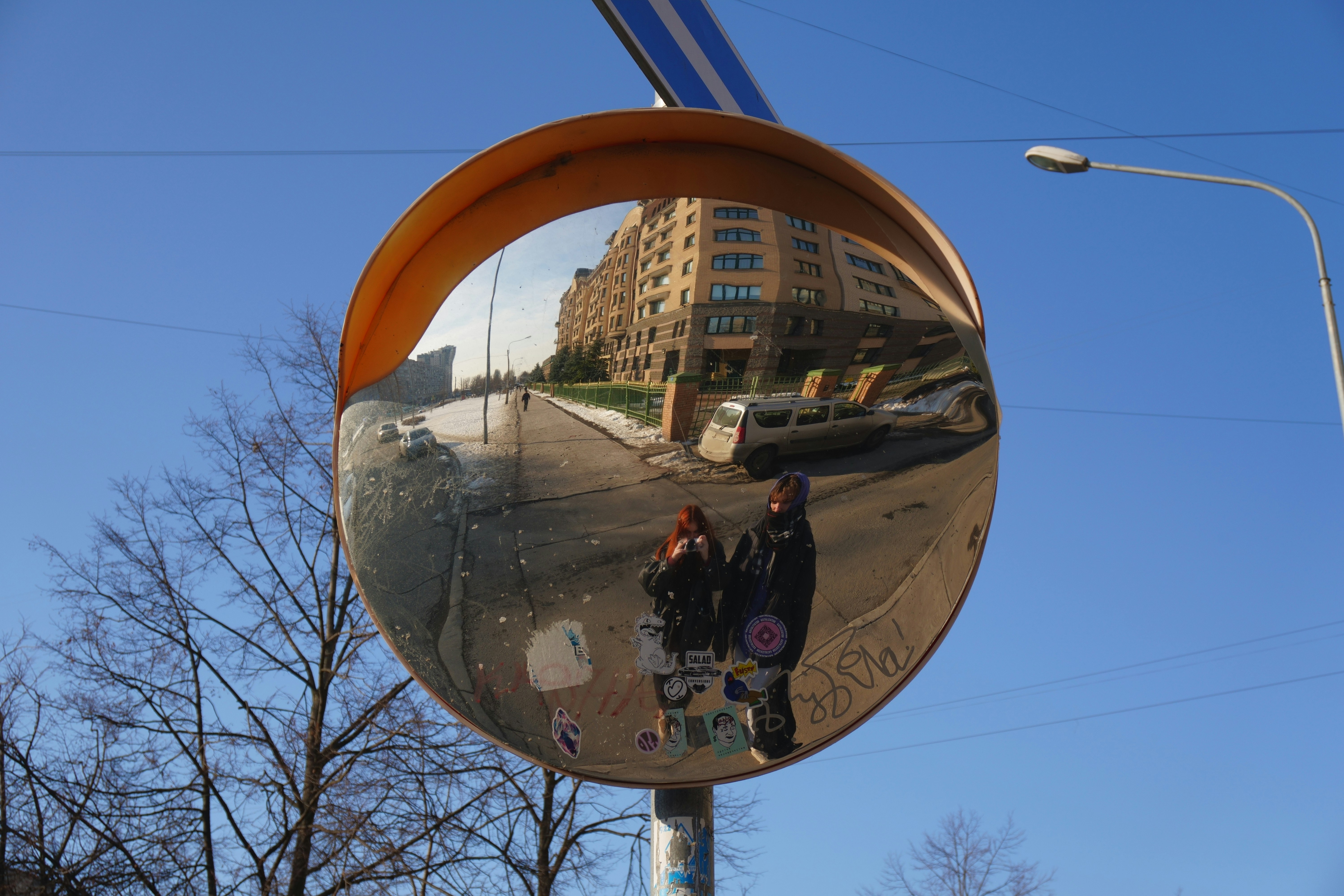A reflective convex mirror capturing the vibrant urban scene and two figures in winter attire, showcasing the interplay of light and surroundings.