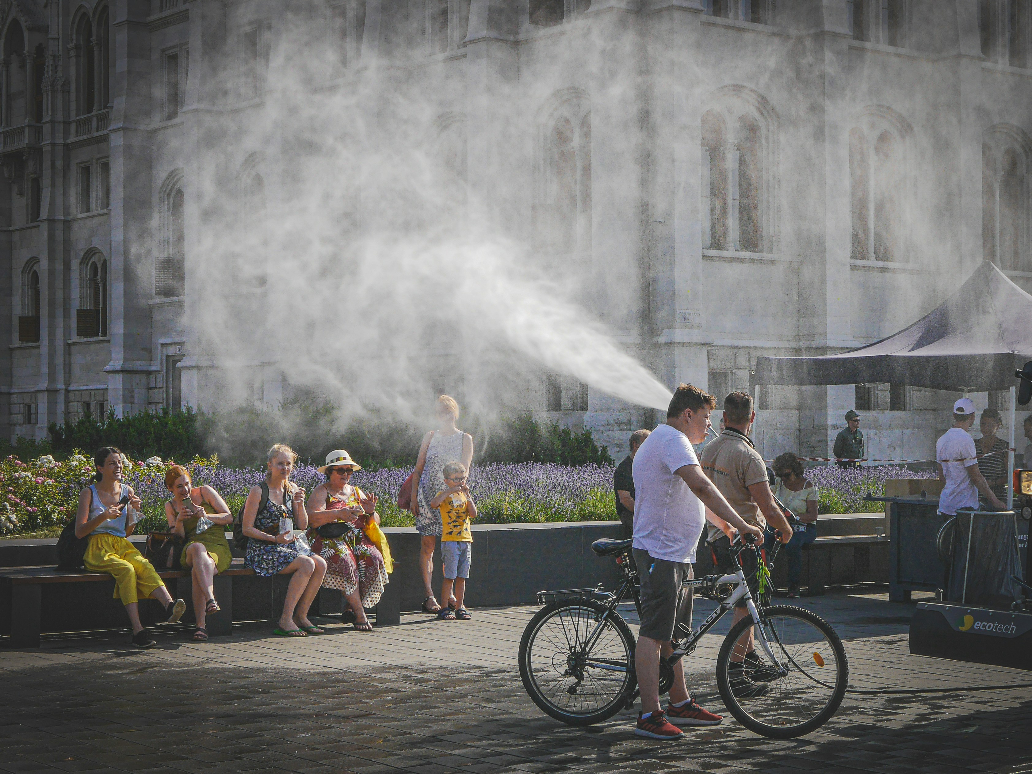 Street scene on a hot summer day in Budapest with a cyclist passing through a cooling mist, people sitting on benches and enjoying the shade. A humorous and refreshing moment of everyday life. | People cooling off from mist spray on a hot day