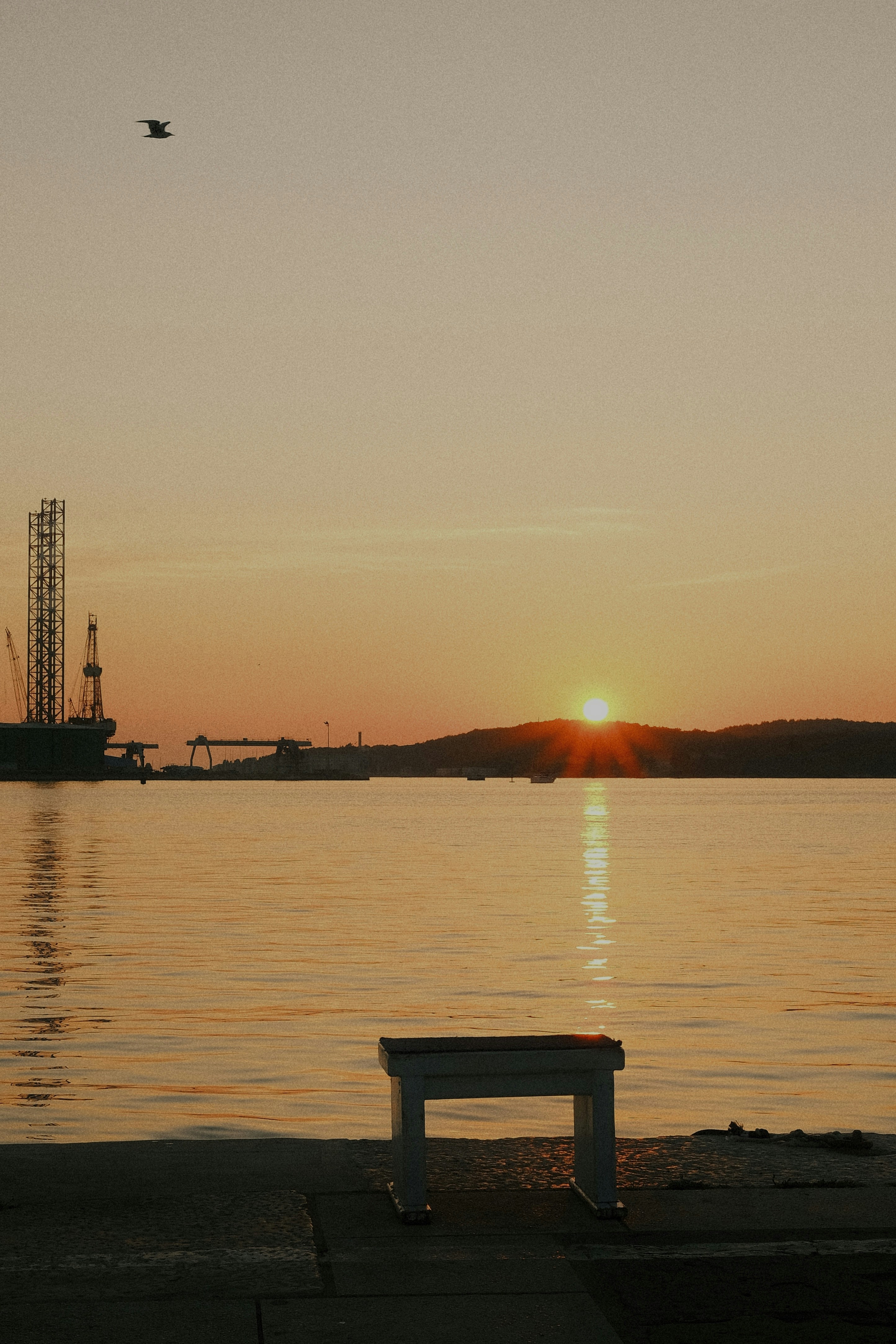 A tranquil scene featuring a bench by the water's edge as the sun sets behind distant hills, casting golden hues over the calm surface. The silhouette of an industrial structure adds contrast to the serene backdrop.
