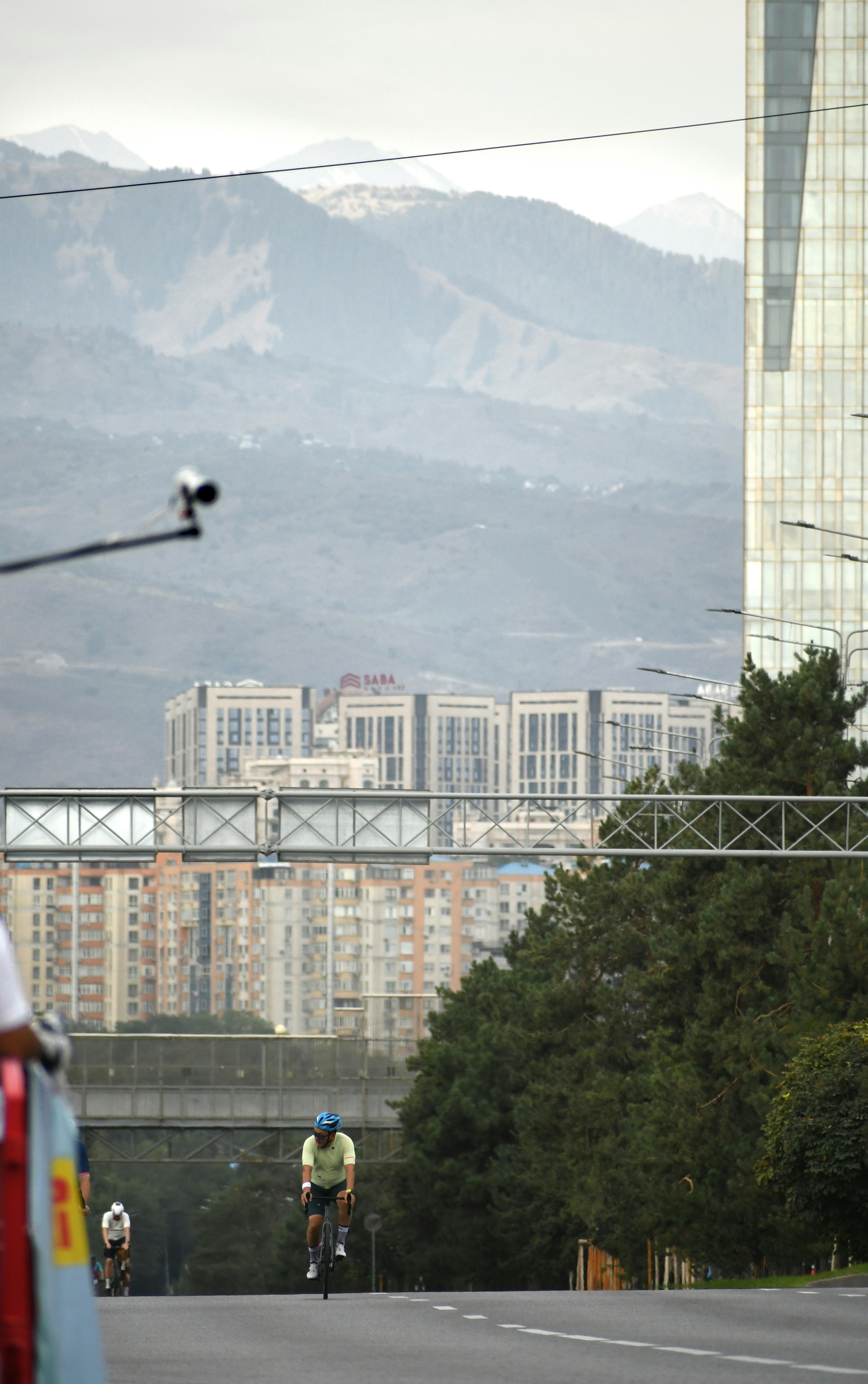 Gran Fondo Almaty 2025 | Cyclists race on a road with city buildings and mountains.