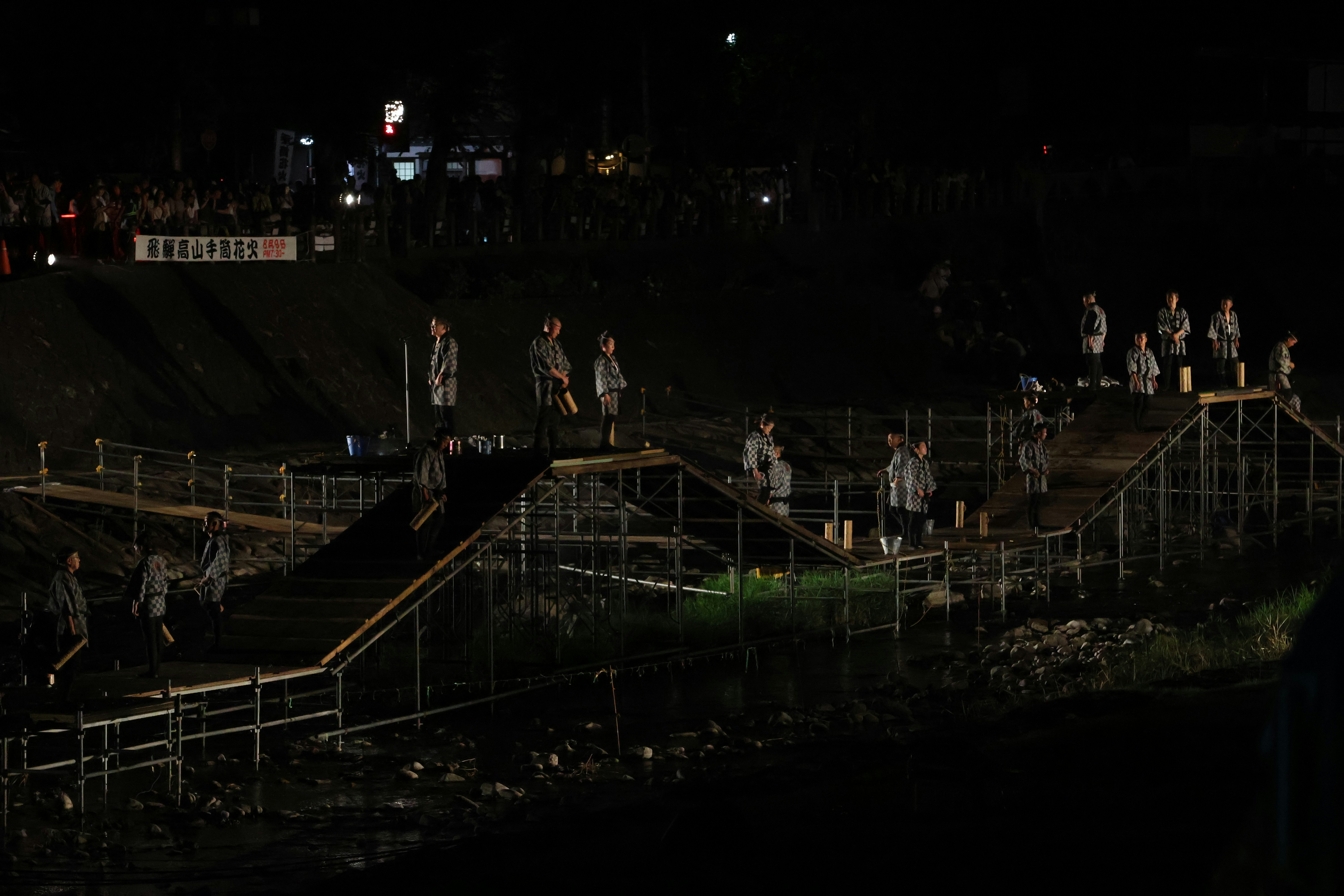 All staff members contributing to the handheld fireworks show lining up and bowing to viewers at the end of the show | People gathered on wooden structures at night