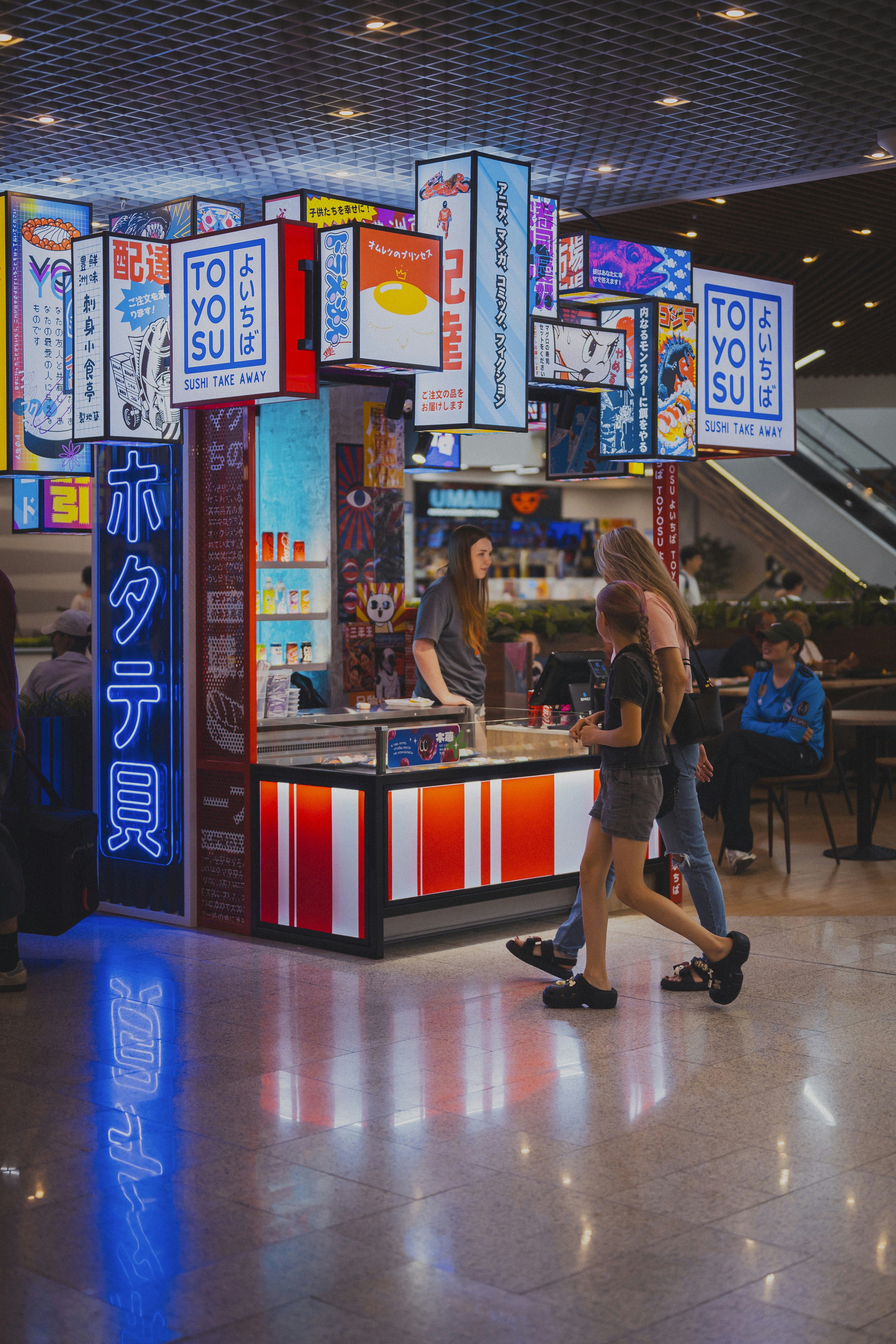 Street food with sushi and rolls in the style of a Japanese metropolis. | People walk past a brightly lit japanese food stall.