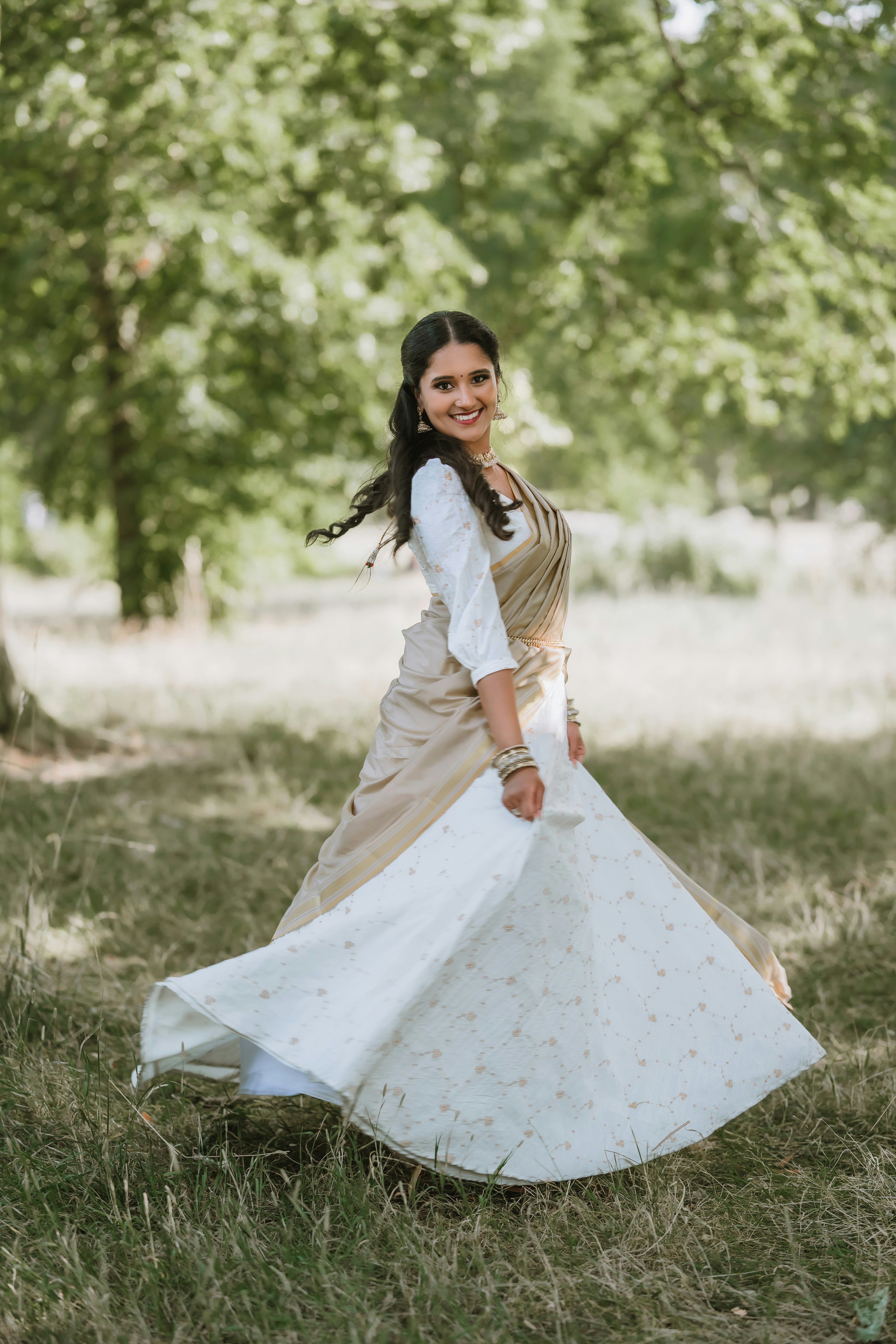 Woman in a flowing traditional outfit twirls gracefully in a sunlit meadow surrounded by lush greenery.