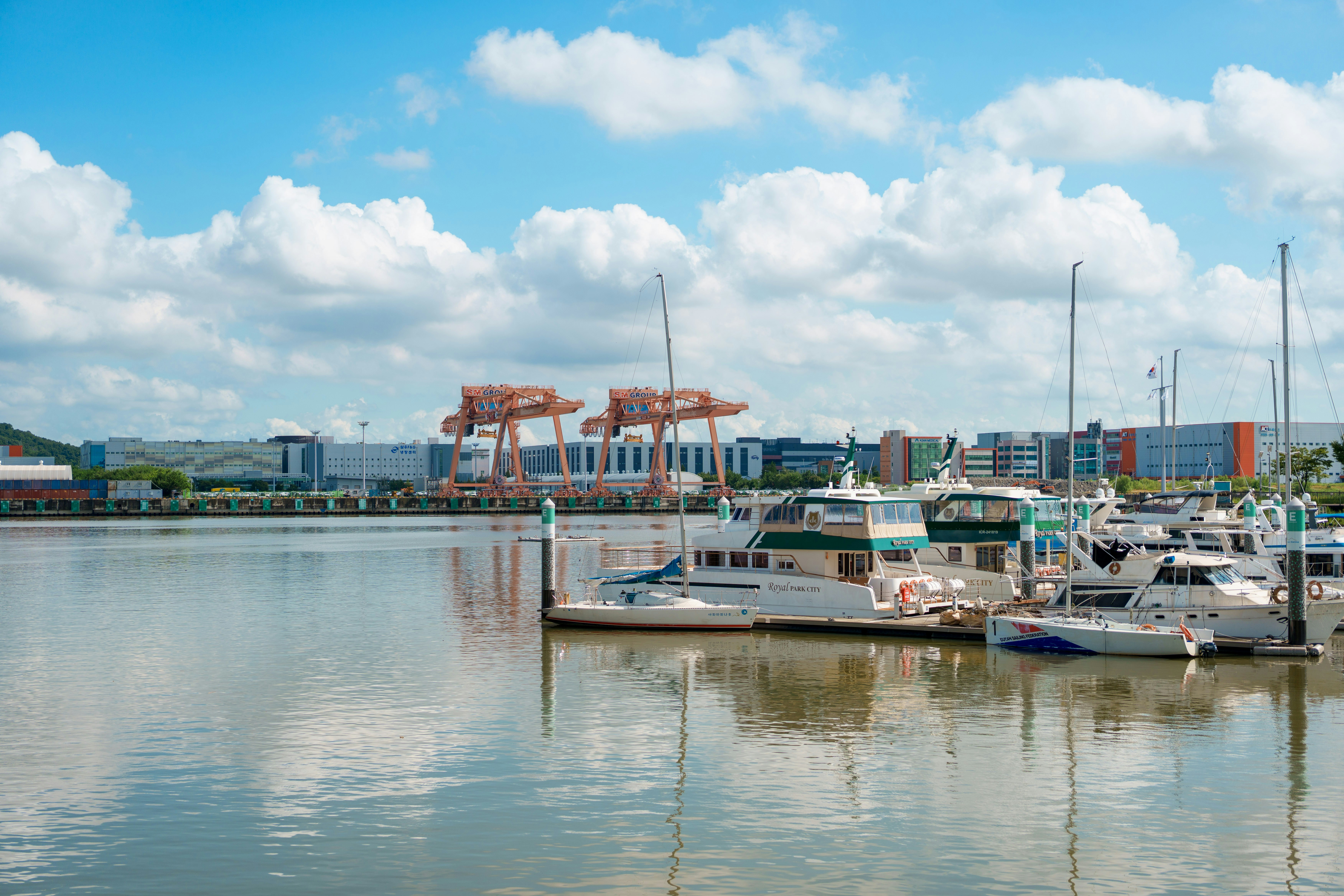 Boats docked in a calm harbor, reflecting the vibrant skyline and industrial cranes under a partly cloudy sky.
