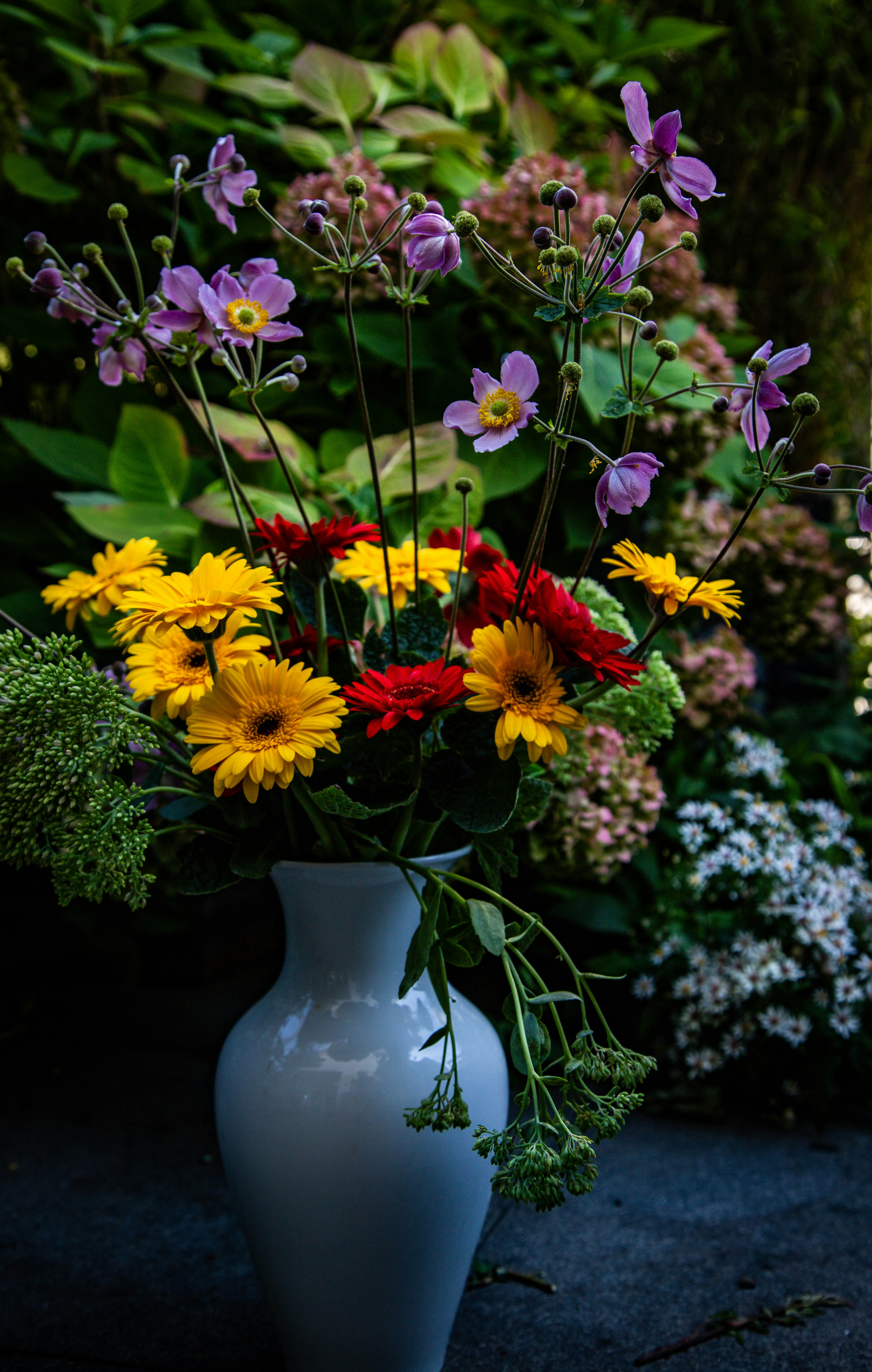 A bouquet of colorful flowers, including yellow gerbera daisies and purple blooms, arranged in a sleek white vase against a lush green backdrop.