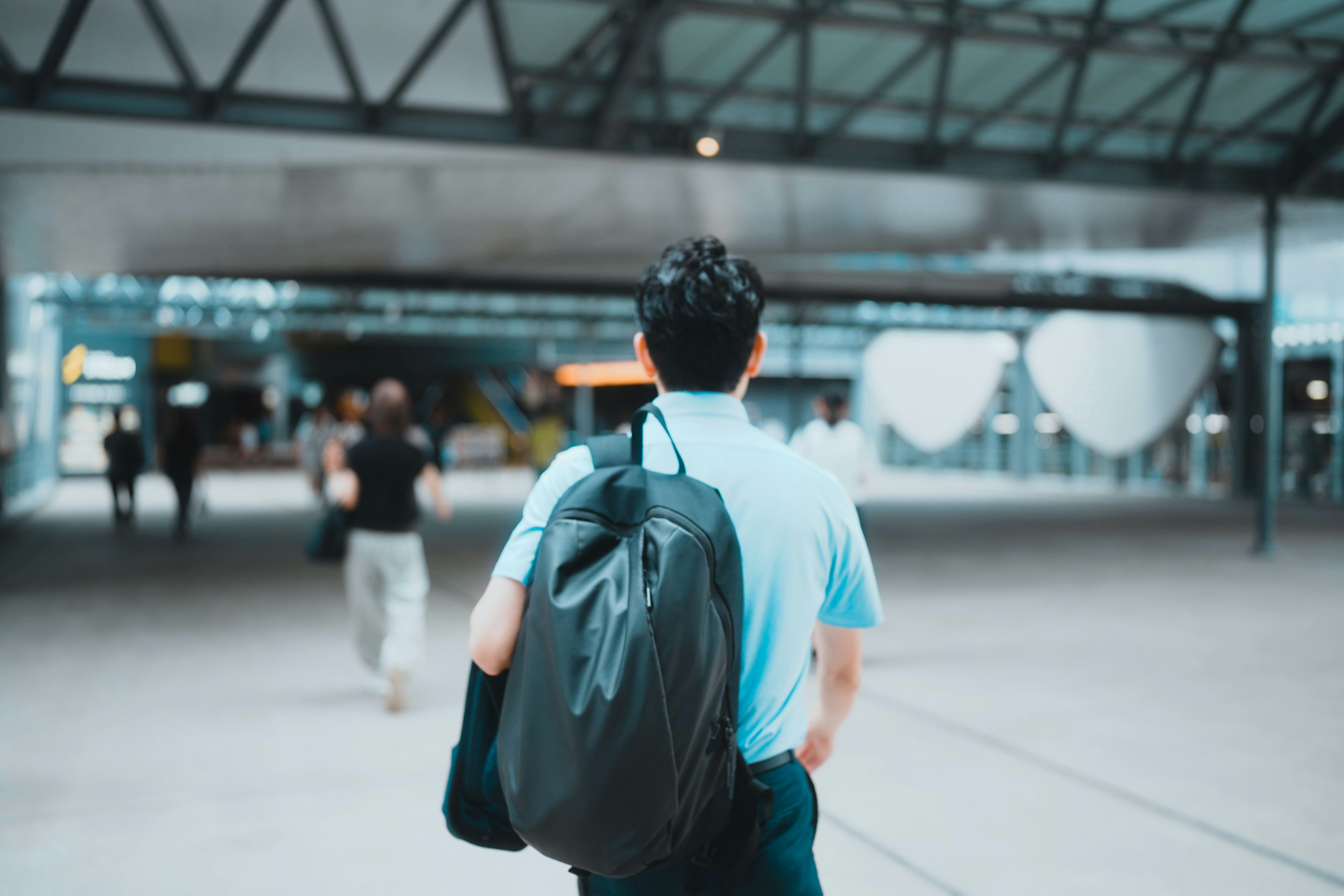 A rear view of a person with a black backpack and a light blue shirt walking away from the camera in a modern, semi-enclosed public space, likely a train station or airport terminal. The background is intentionally blurred, with other people and a metal, industrial-style ceiling visible, creating a sense of motion and daily life. | Man with backpack walks through modern building.