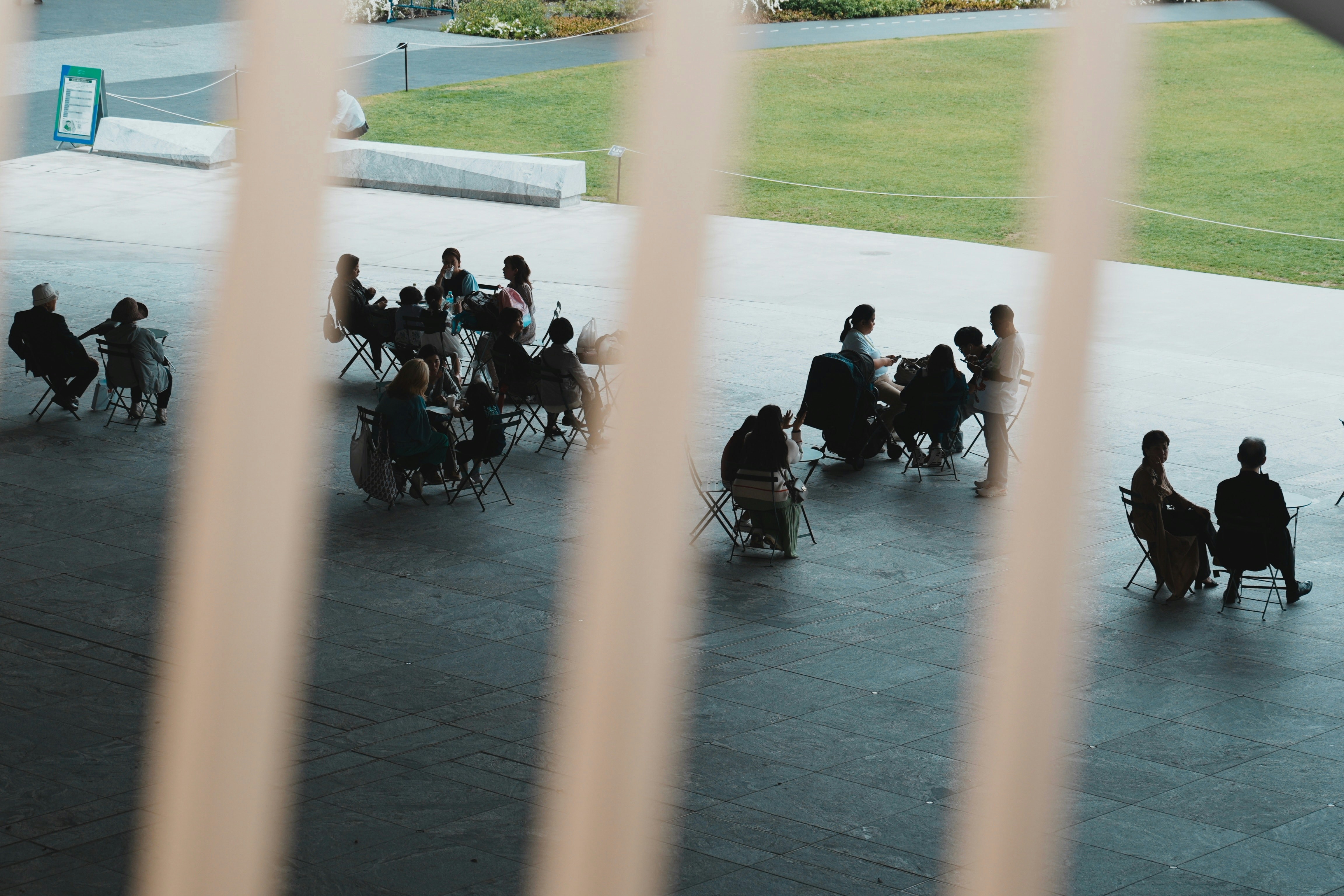 An elevated, somewhat obscured view of several groups of people sitting at small tables and chairs in an outdoor public space. The image is framed by a blurry, light-colored structure in the foreground, with a grassy area and paved ground visible in the background. The scene depicts a casual, social gathering, and has a soft, moody aesthetic. | People sitting at tables outside behind bars.