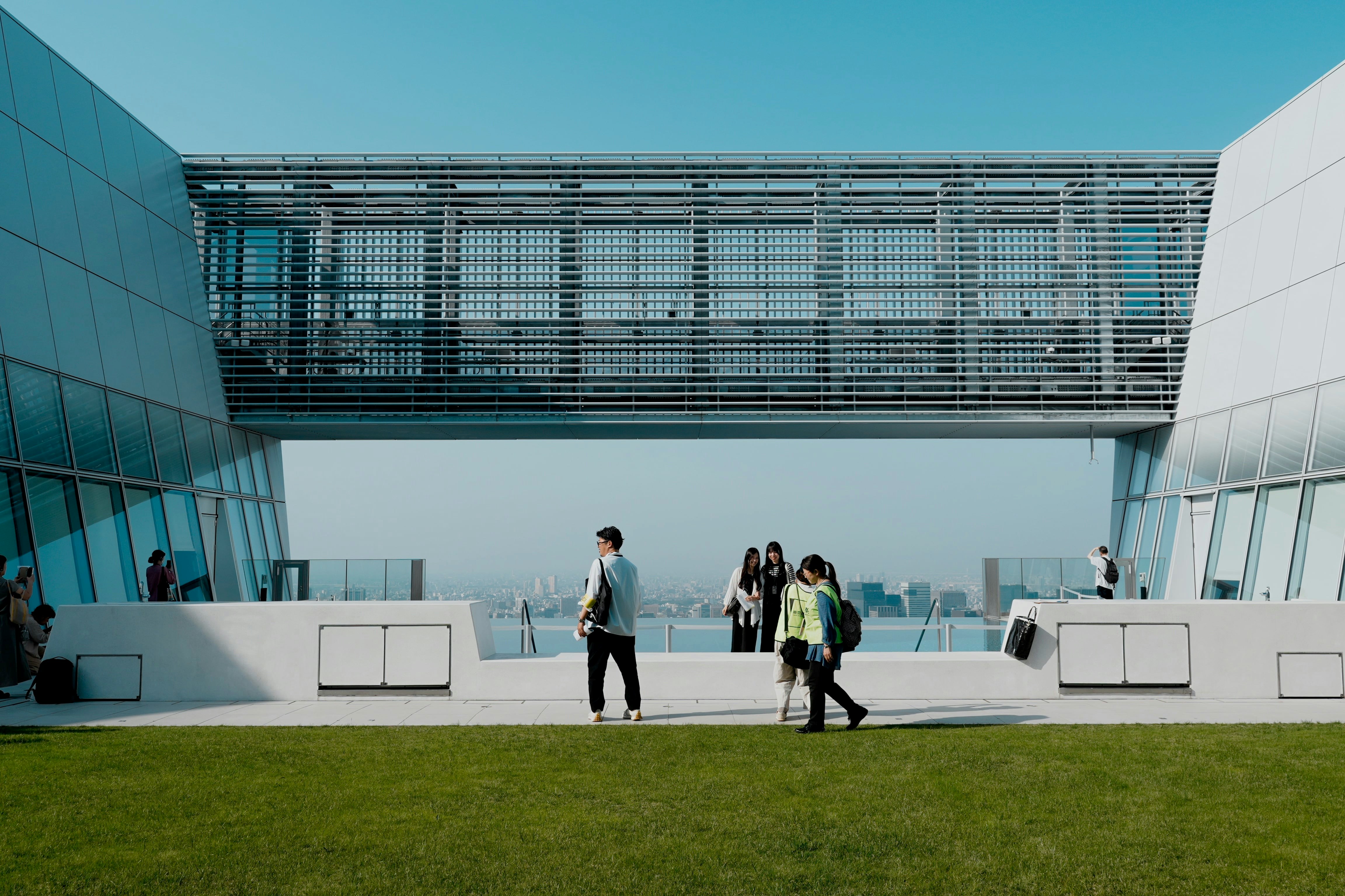 People walking across a green lawn beneath a sleek architectural structure with a grid design, showcasing a blend of nature and modernity.