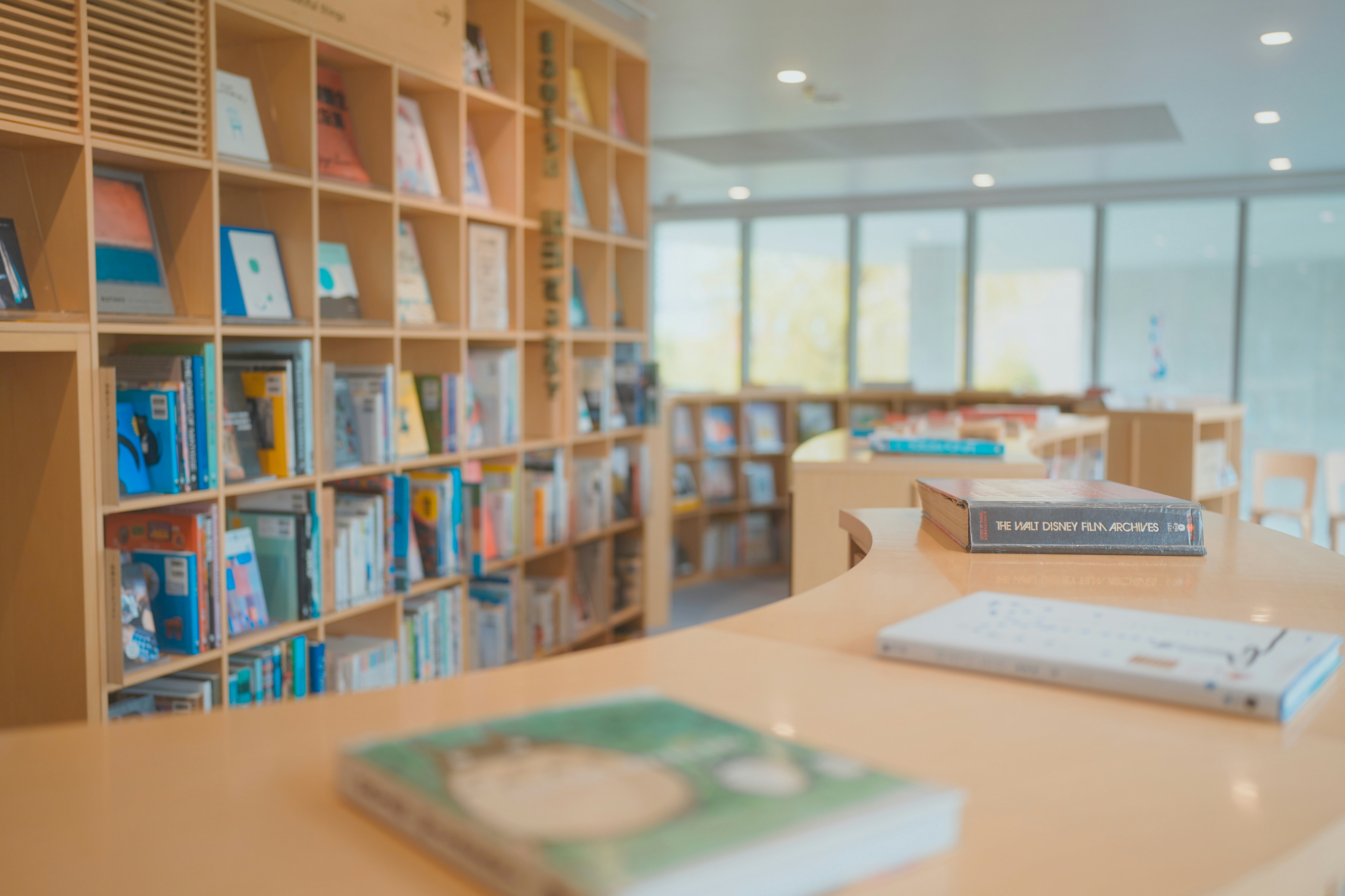 A soft-focus, interior shot of a modern and bright library or bookstore. The scene features wooden bookshelves filled with various books on the left, a curved counter with a few books placed on top in the foreground, and large windows that let in natural light. | Books on shelves in a modern library reading area.