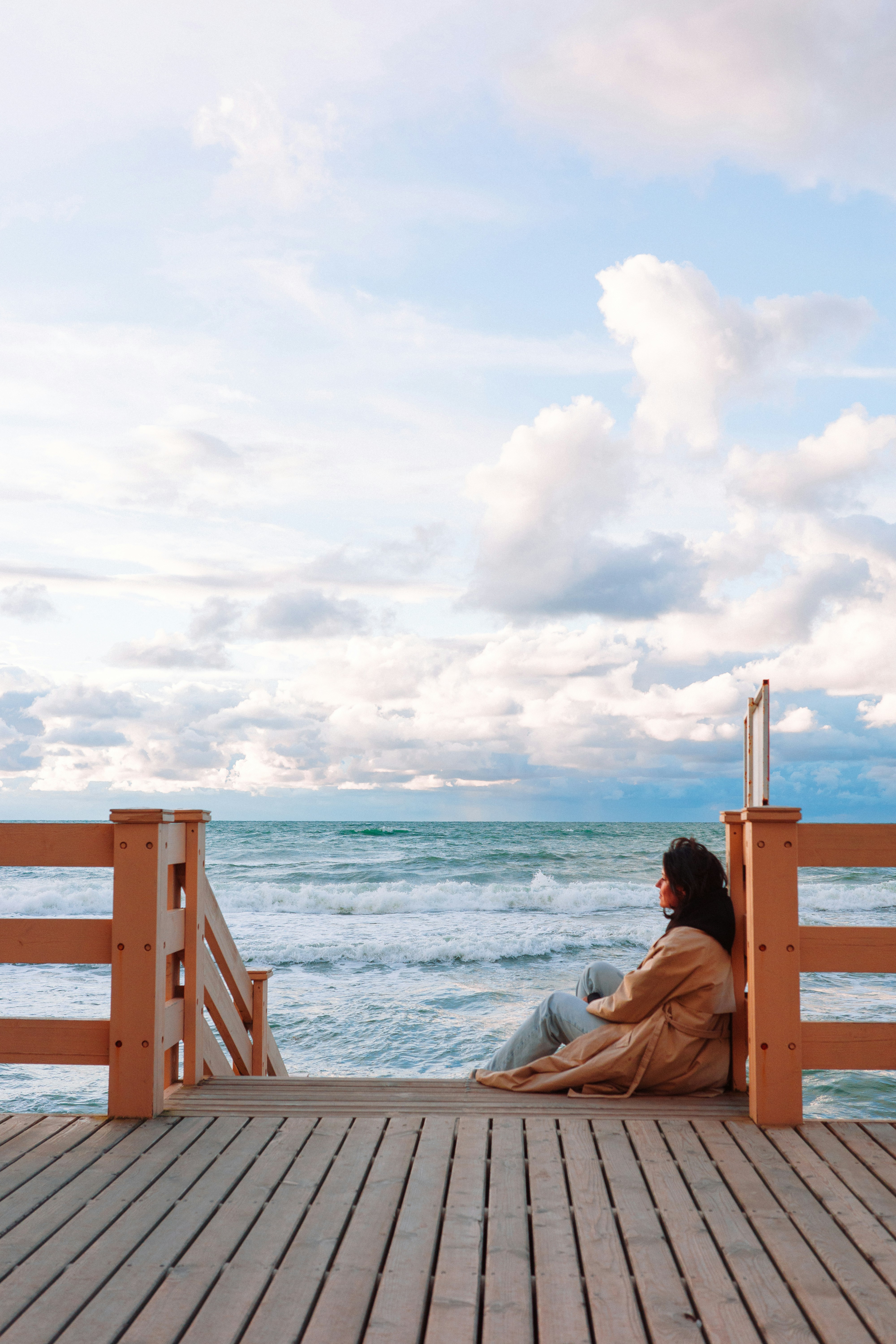A person seated on a wooden pier gazes thoughtfully at the ocean under a sky filled with fluffy clouds.