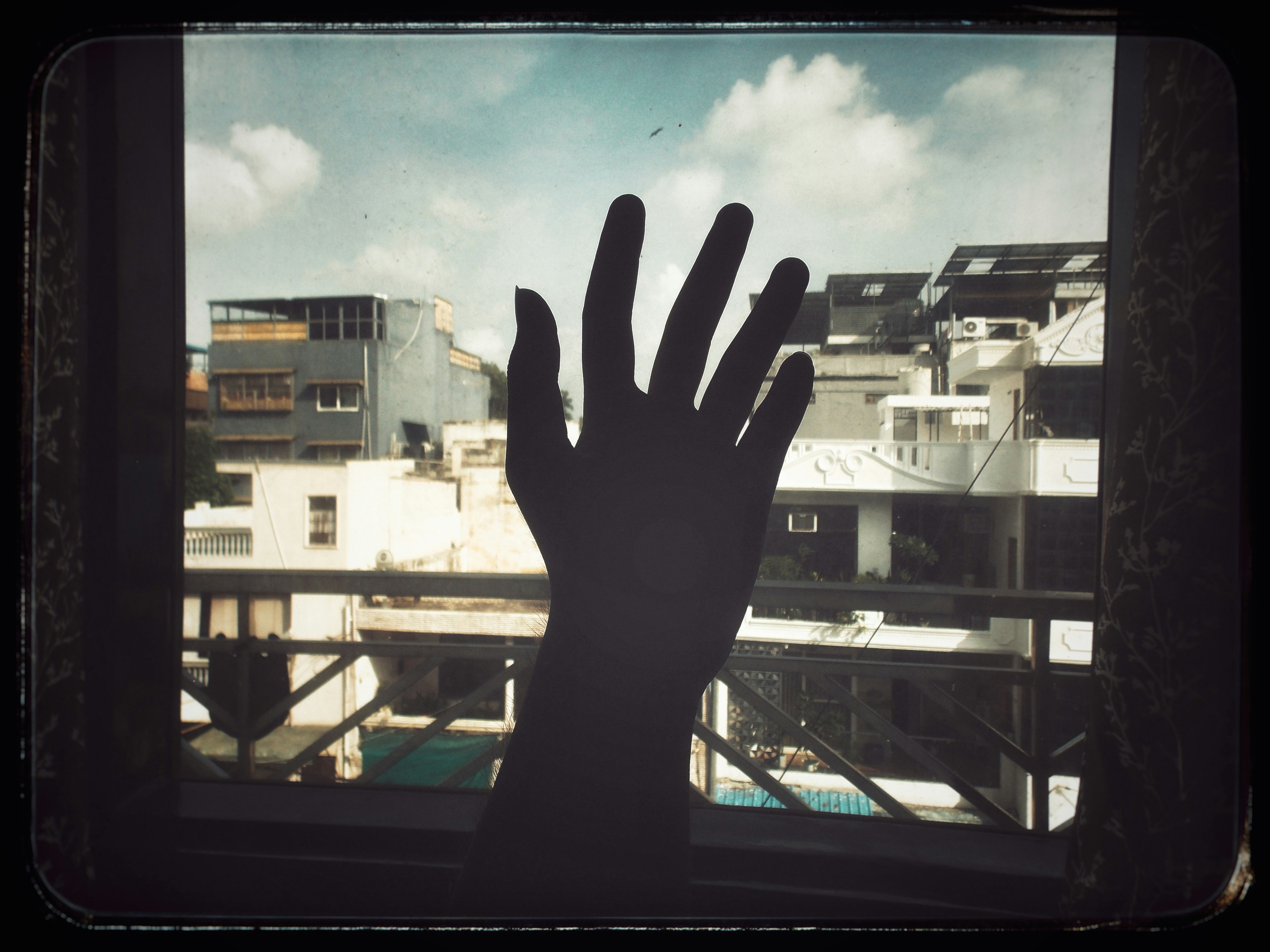 A silhouette of a hand raised against the backdrop of an urban skyline and cloudy blue sky. Symbolic, artistic, and minimal. | Silhouette of a hand waving from a window