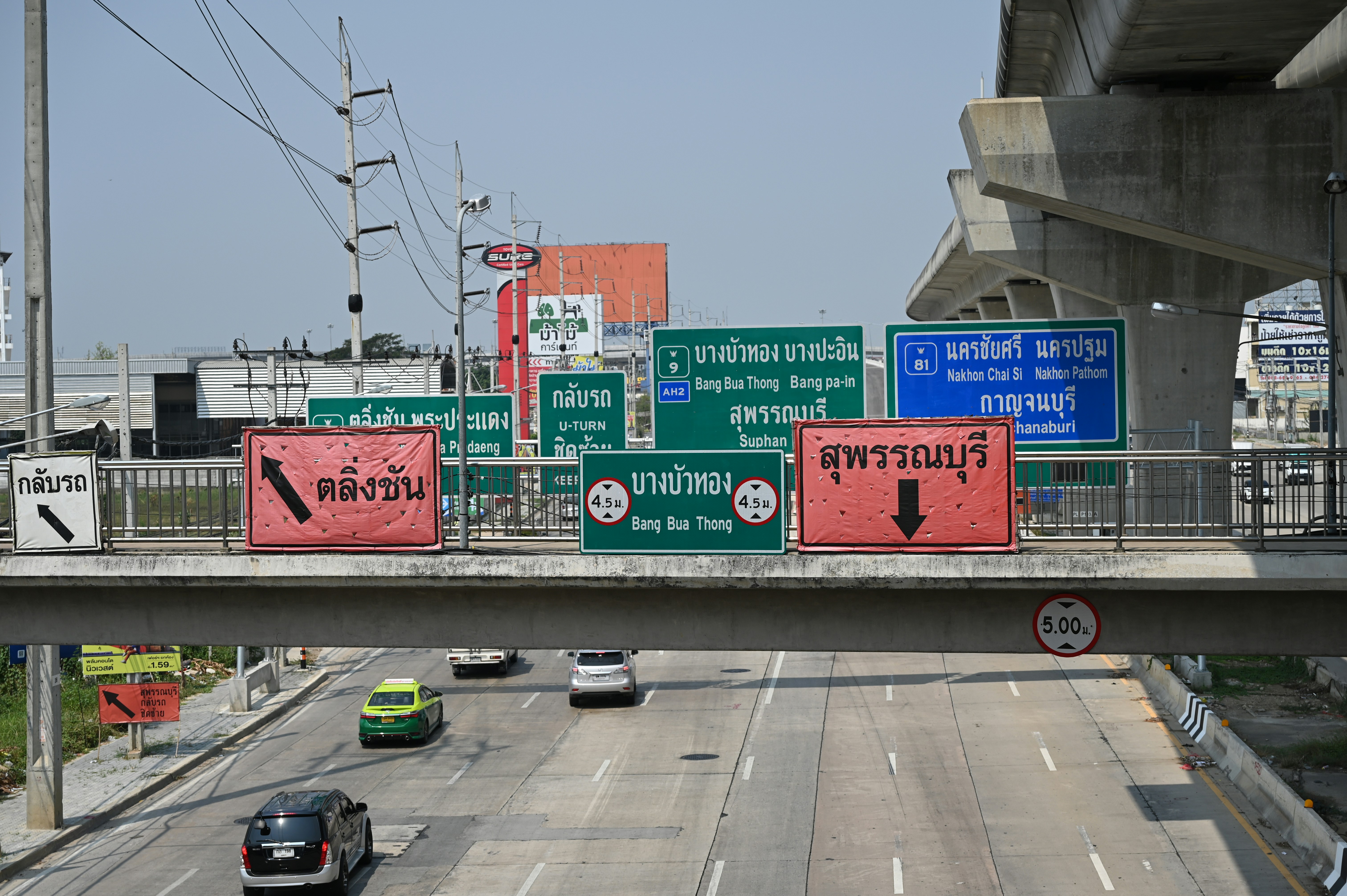 A collection of road signs directing traffic in Bangkok, showcasing various routes and destinations. The signs are prominently displayed above a busy street.