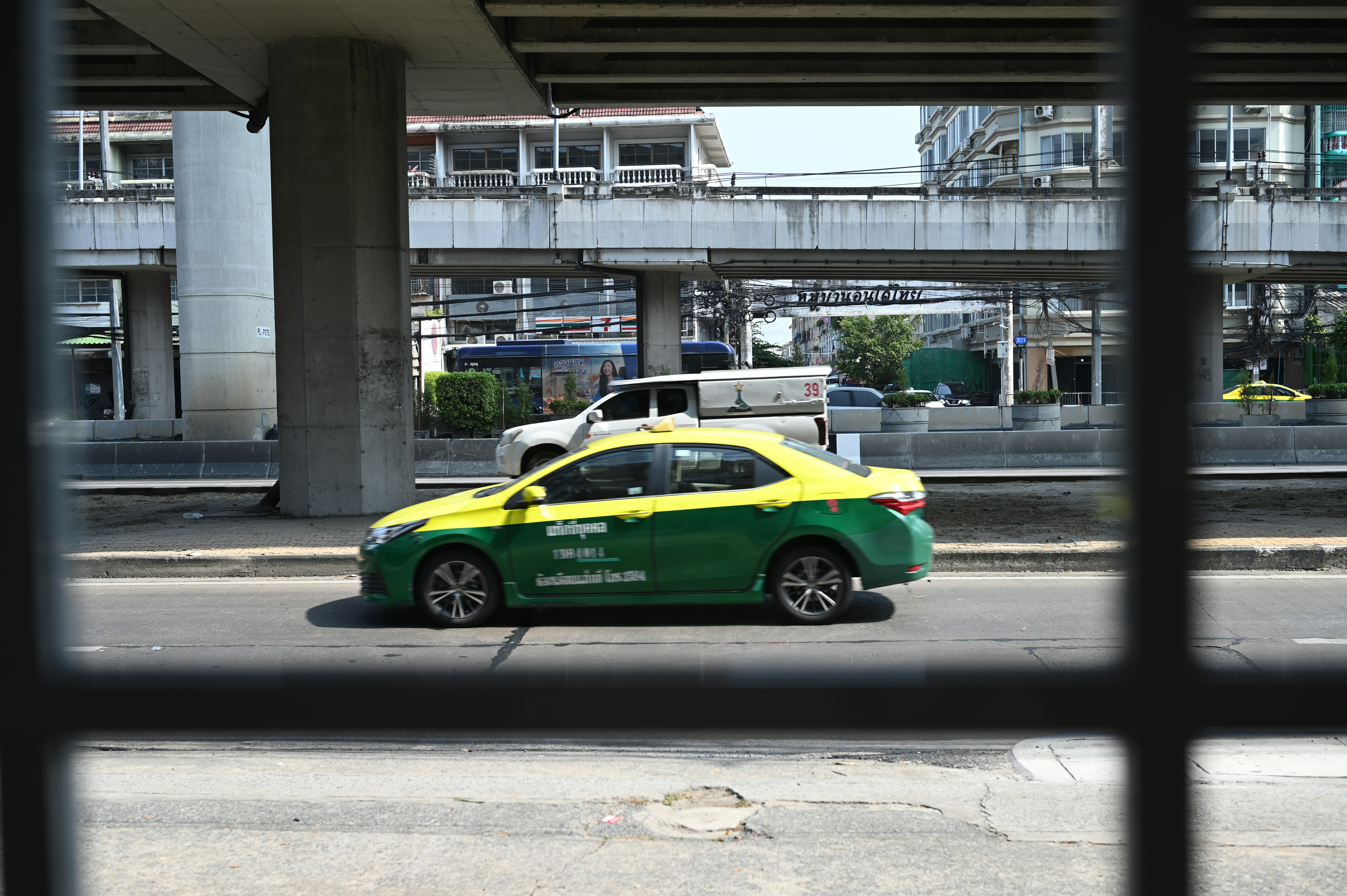 Green and yellow taxi navigating city streets, framed by a grid of metal bars. Urban infrastructure looms in the background.