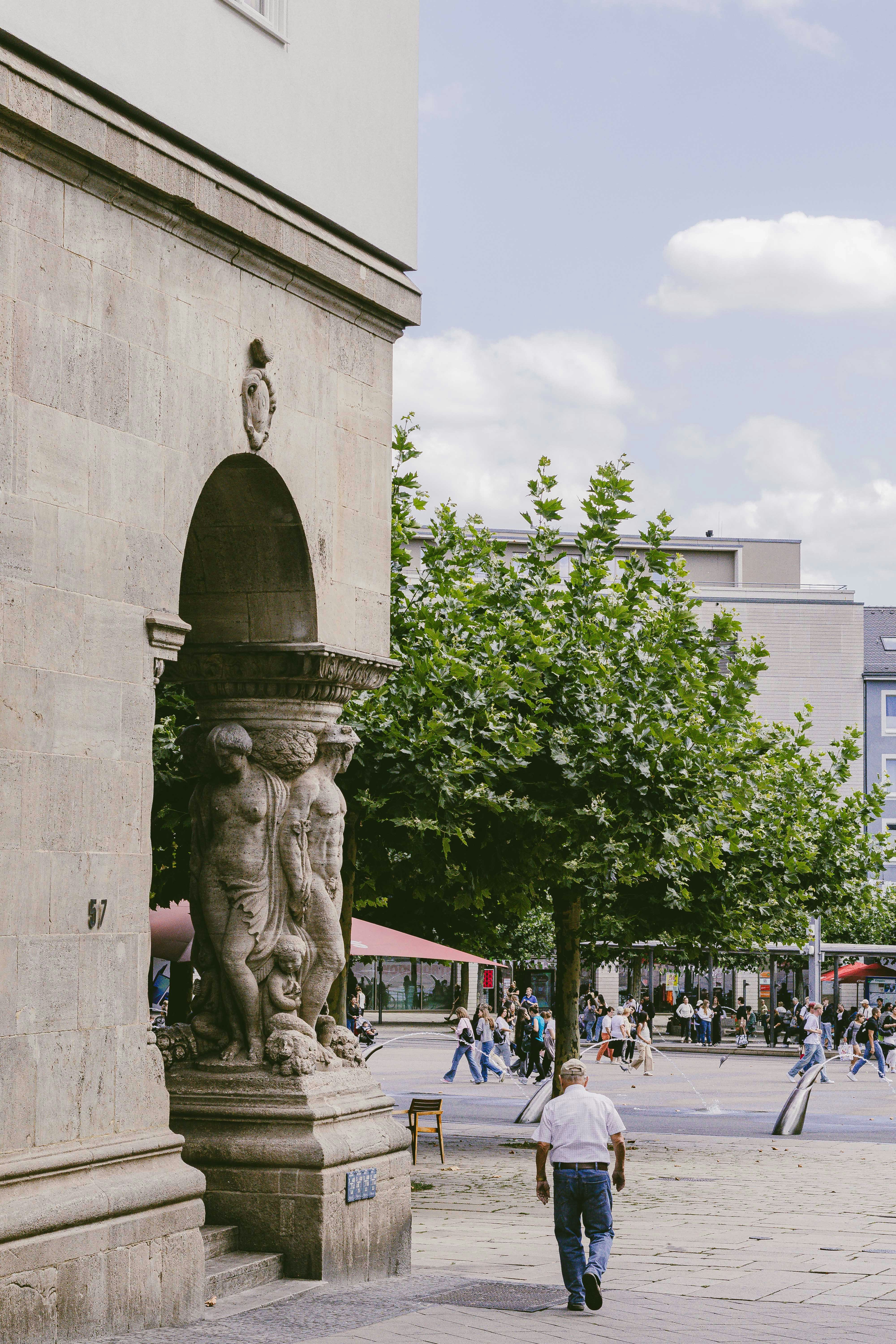 Intricate stone sculptures frame a lively plaza, where pedestrians traverse under the shade of lush trees. A blend of architectural elegance and urban life.