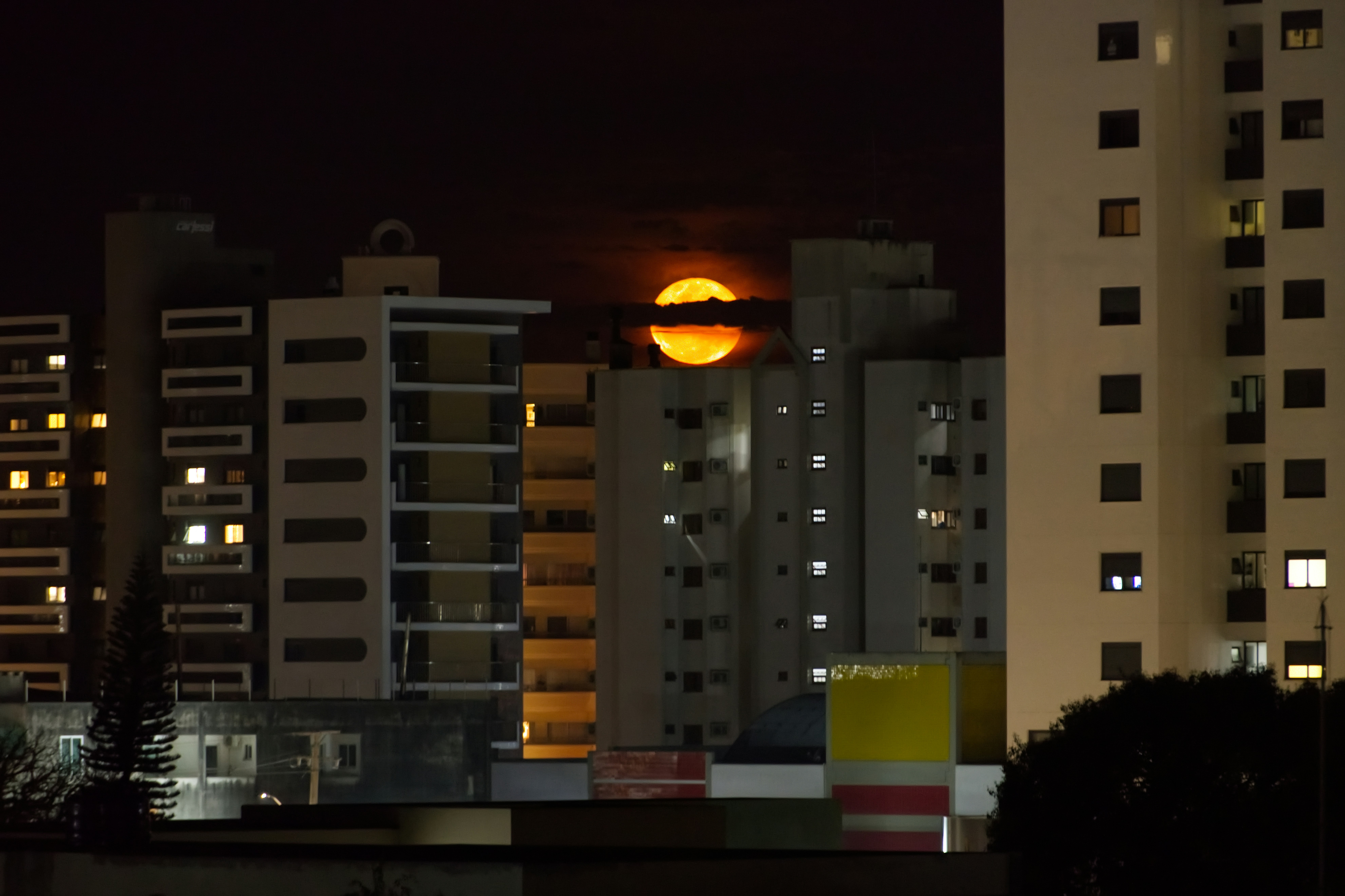 The sun sets dramatically behind urban buildings, casting a warm glow amidst the cool night. The scene captures the contrast between natural beauty and architectural lines.
