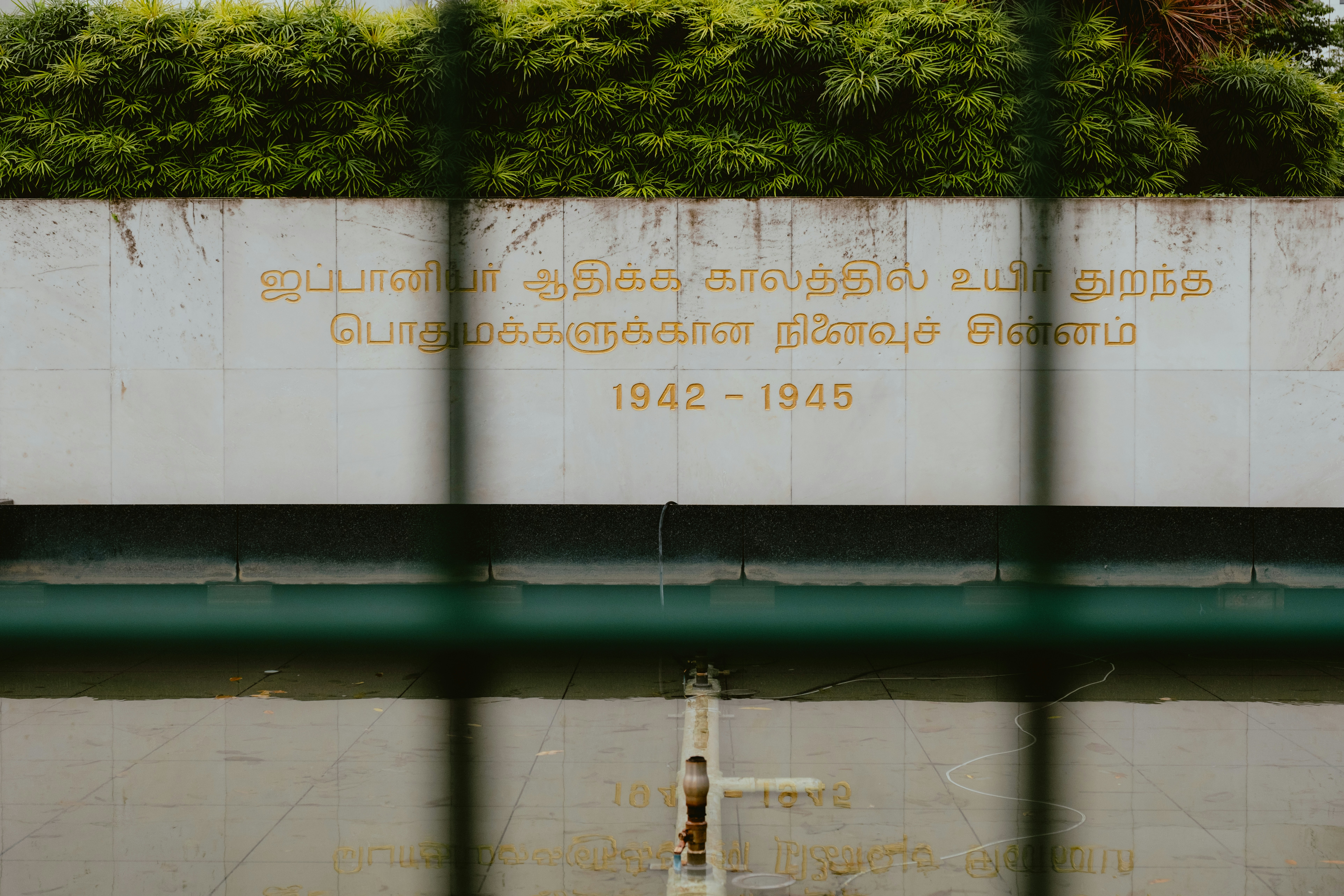 A memorial plaque inscribed with dates and text, partially obscured by a fence, surrounded by lush greenery.