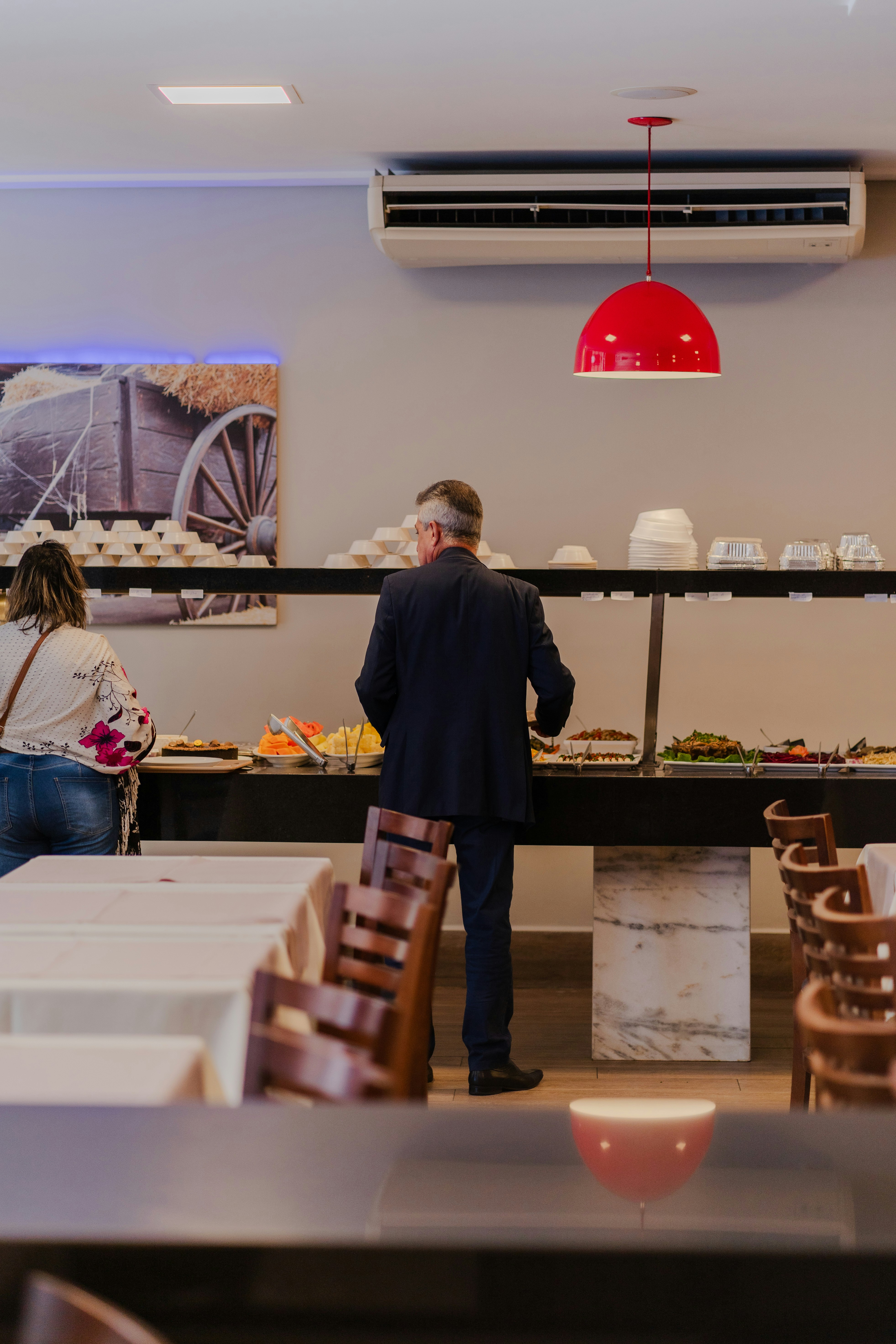 A man in a suit surveys a buffet spread in a modern dining area, illuminated by a striking red pendant light. Various dishes are presented on a long table against a backdrop of a rustic-themed wall.