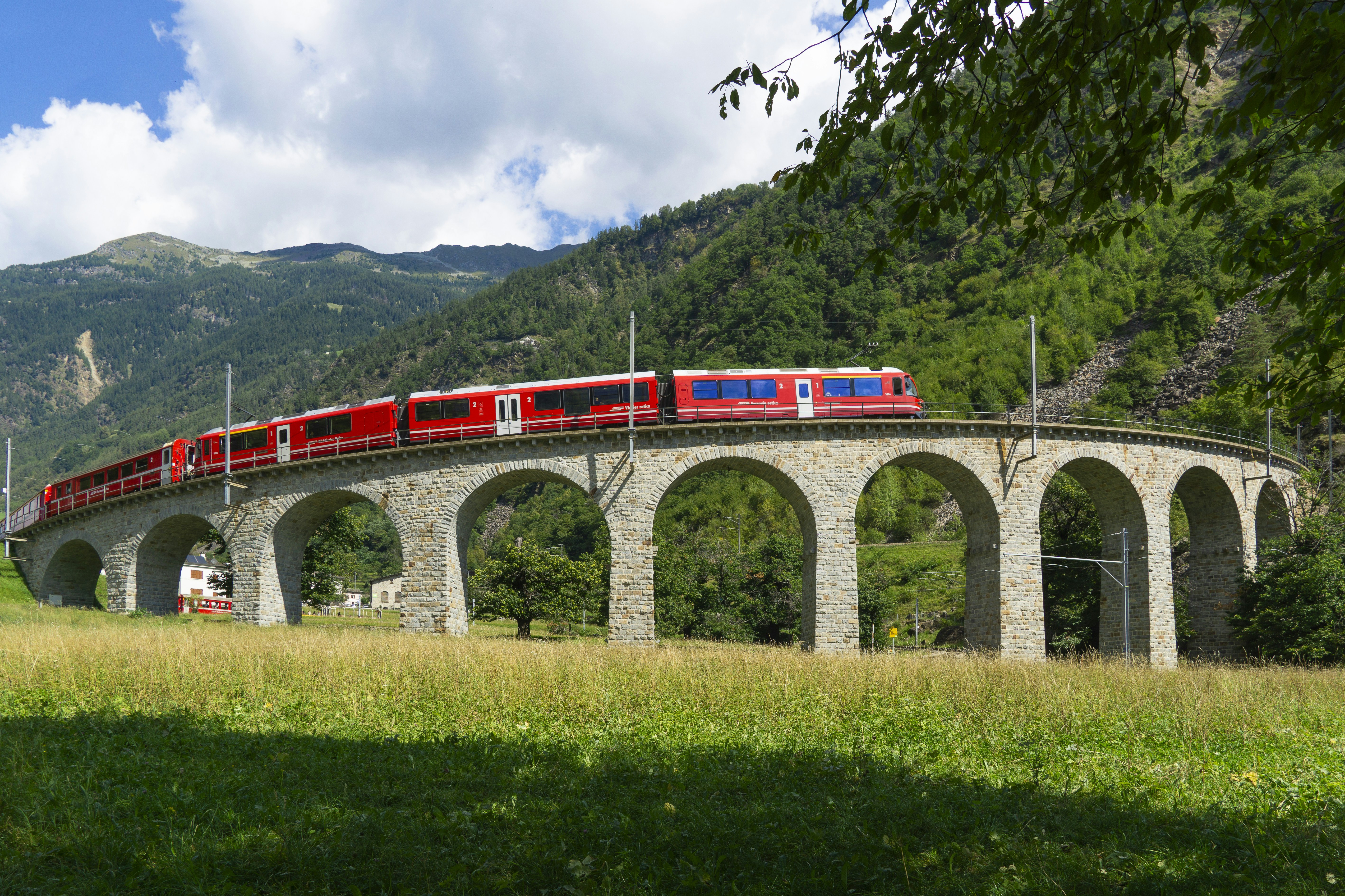 Red train crosses a stone viaduct in the mountains.