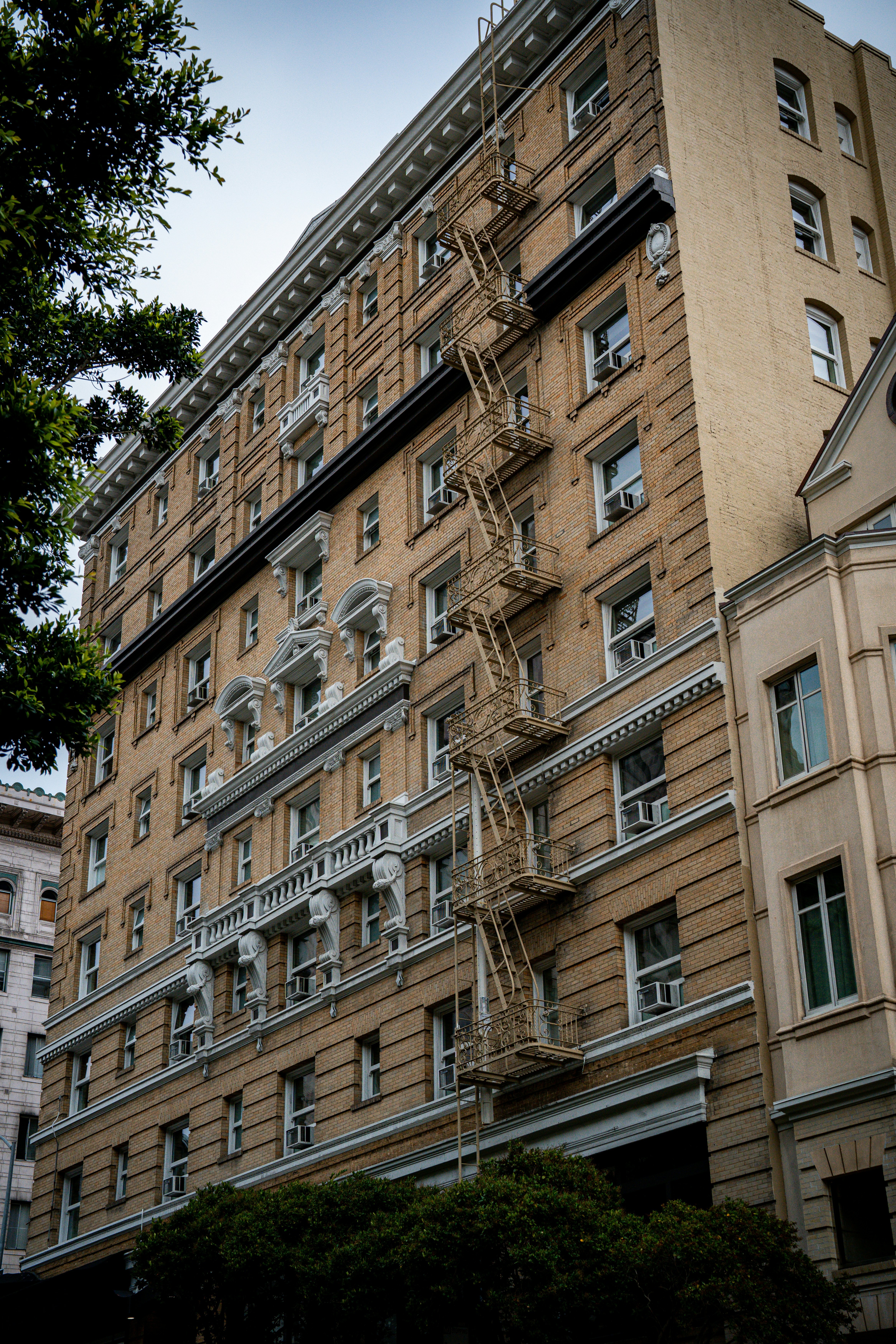A historic building featuring intricate architectural details and a prominent fire escape, framed by lush greenery. The structure showcases a blend of classic design and urban life.