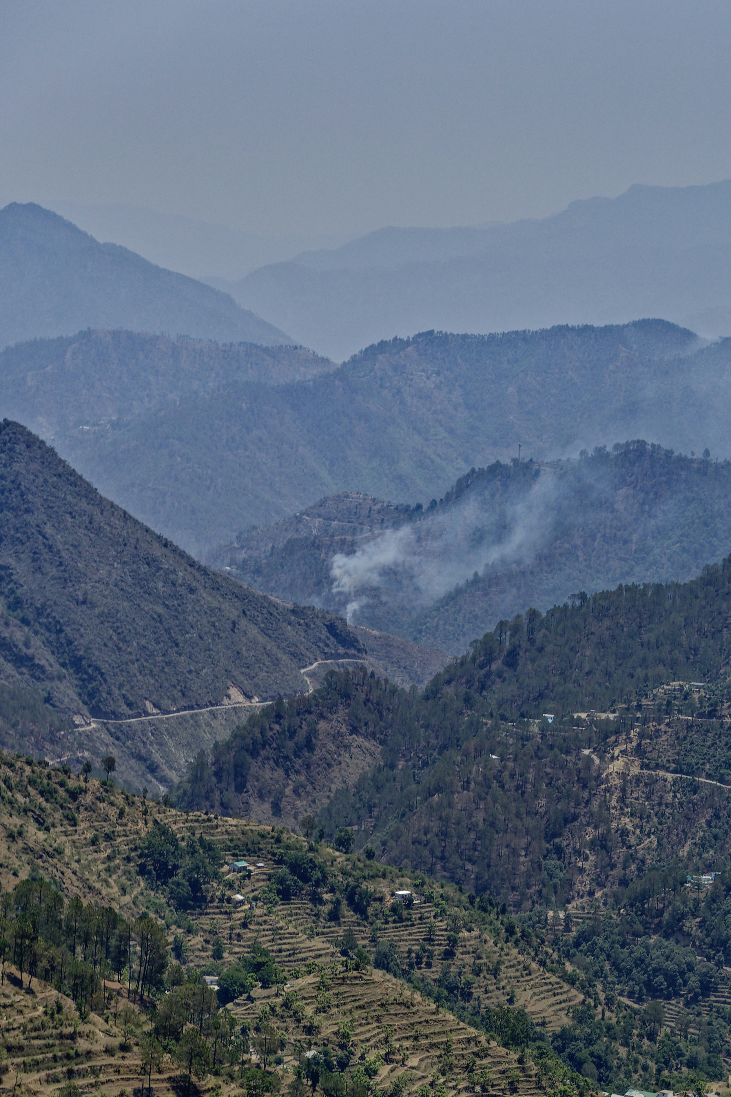 Layered blue mountains with a winding road and smoke.