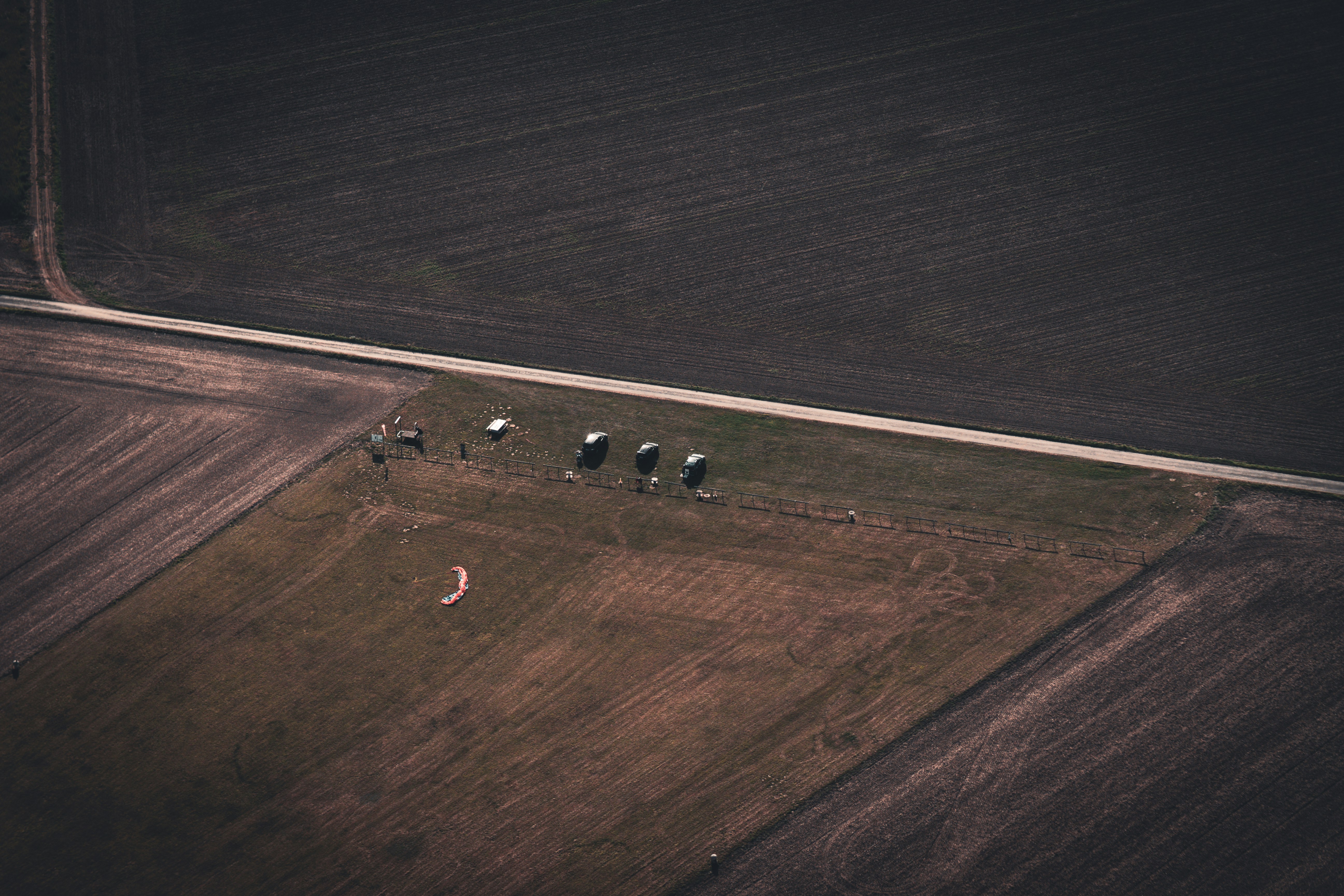 Paragliding field seen from above with cars parked nearby and a colorful wing lying on the ground. Aerial perspective capturing the quiet atmosphere of rural aviation. | Paraglider flying over rural fields near buildings