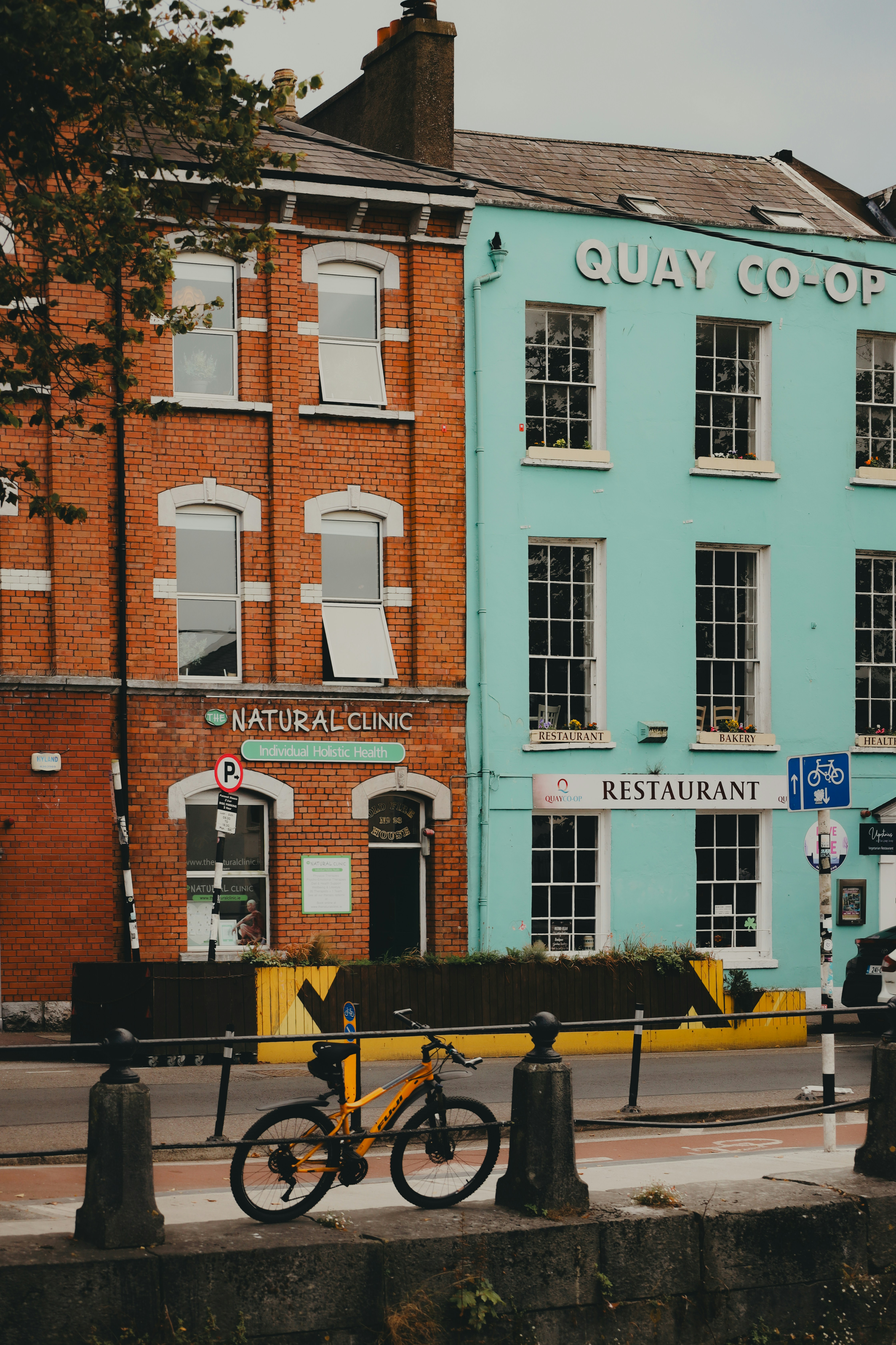 Vibrant buildings line a street by the water, featuring a natural clinic and a restaurant, with a bicycle parked in the foreground.