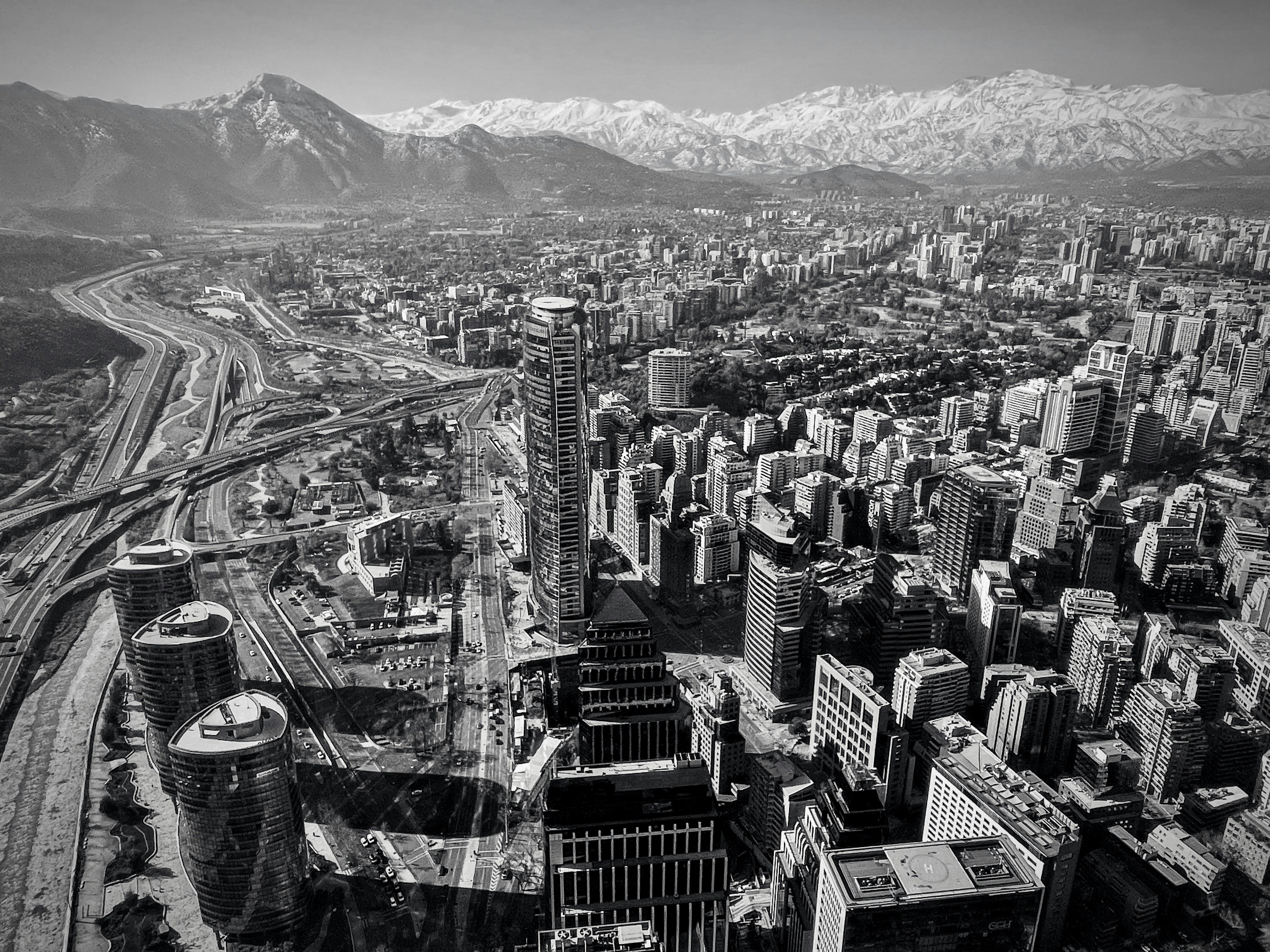 Aerial view of a bustling cityscape juxtaposed with snow-capped mountains in the background, captured in monochrome.