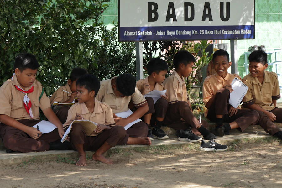 Group of schoolchildren seated outside, engaged in their studies while surrounded by nature and a school sign. 