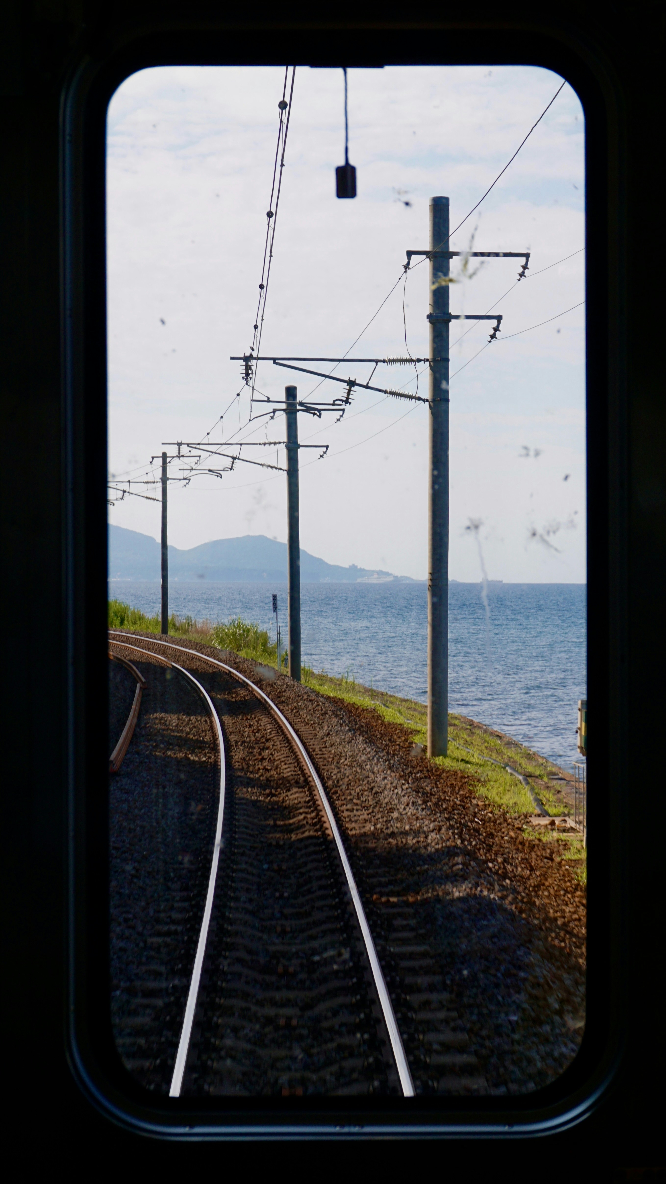 Train tracks curve along the ocean with overhead wires.