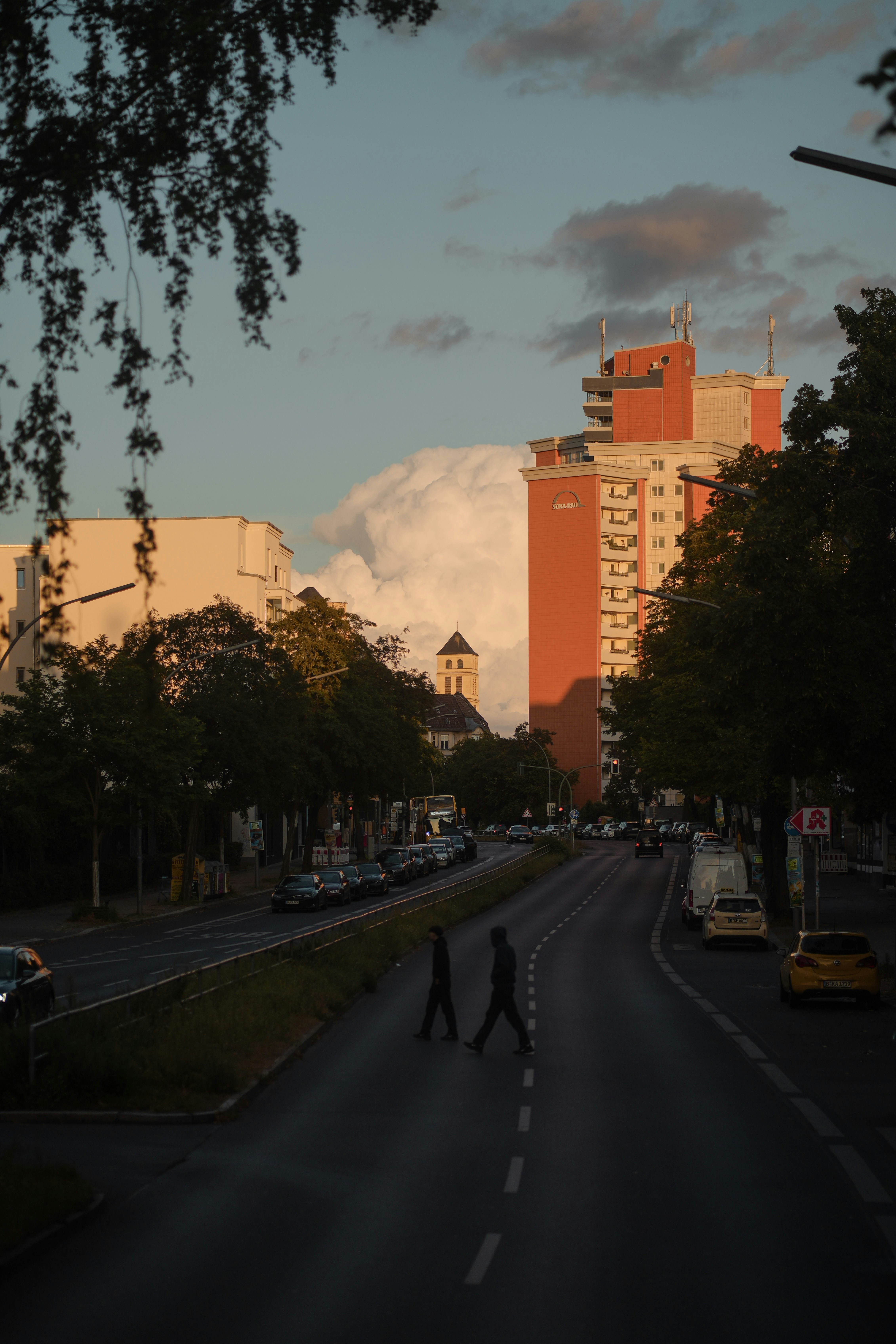 Two figures cross a street in a city at sunset.