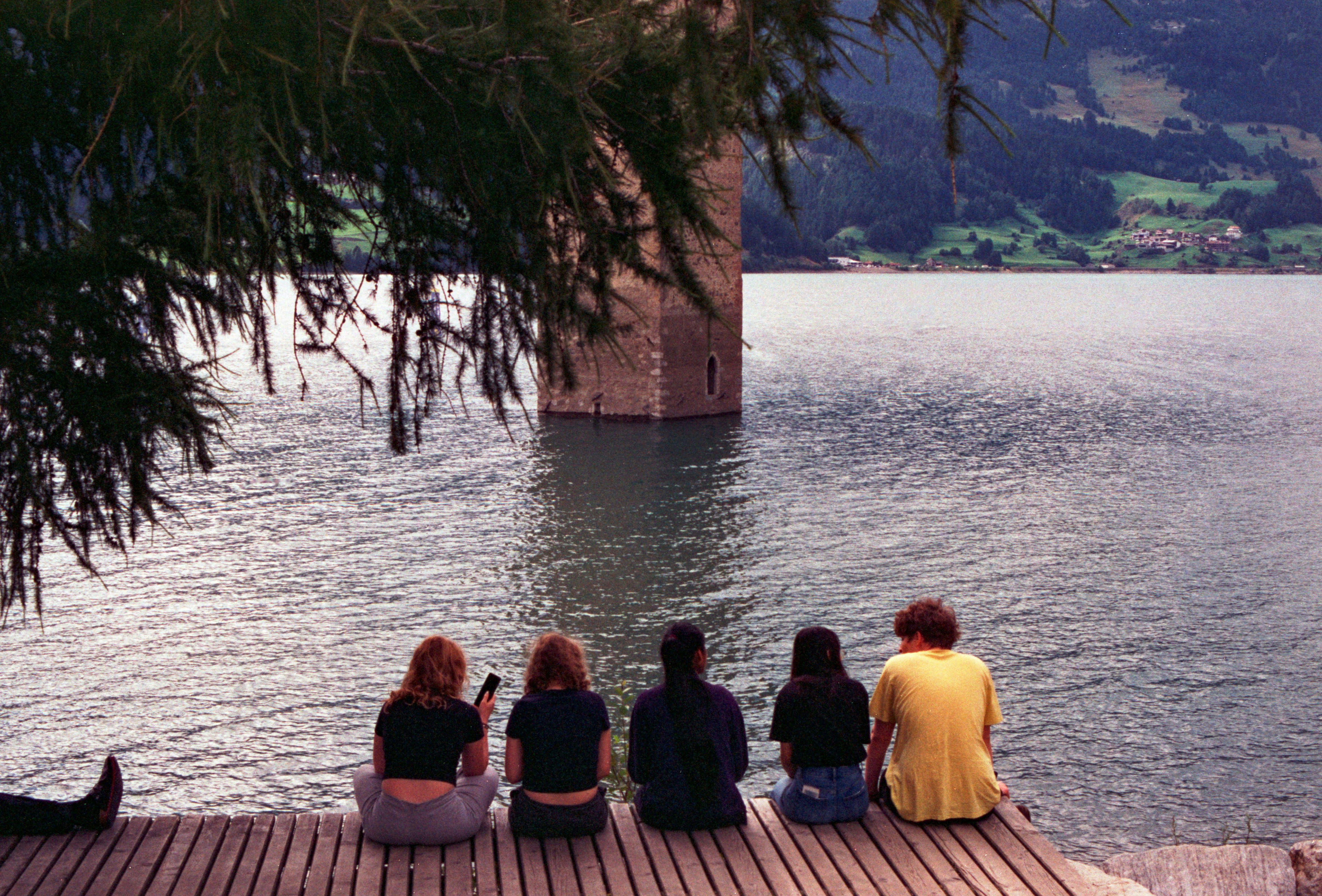 Five friends seated on a wooden dock, gazing out over a serene lake with a distant tower. The scene captures a moment of connection and tranquility.
