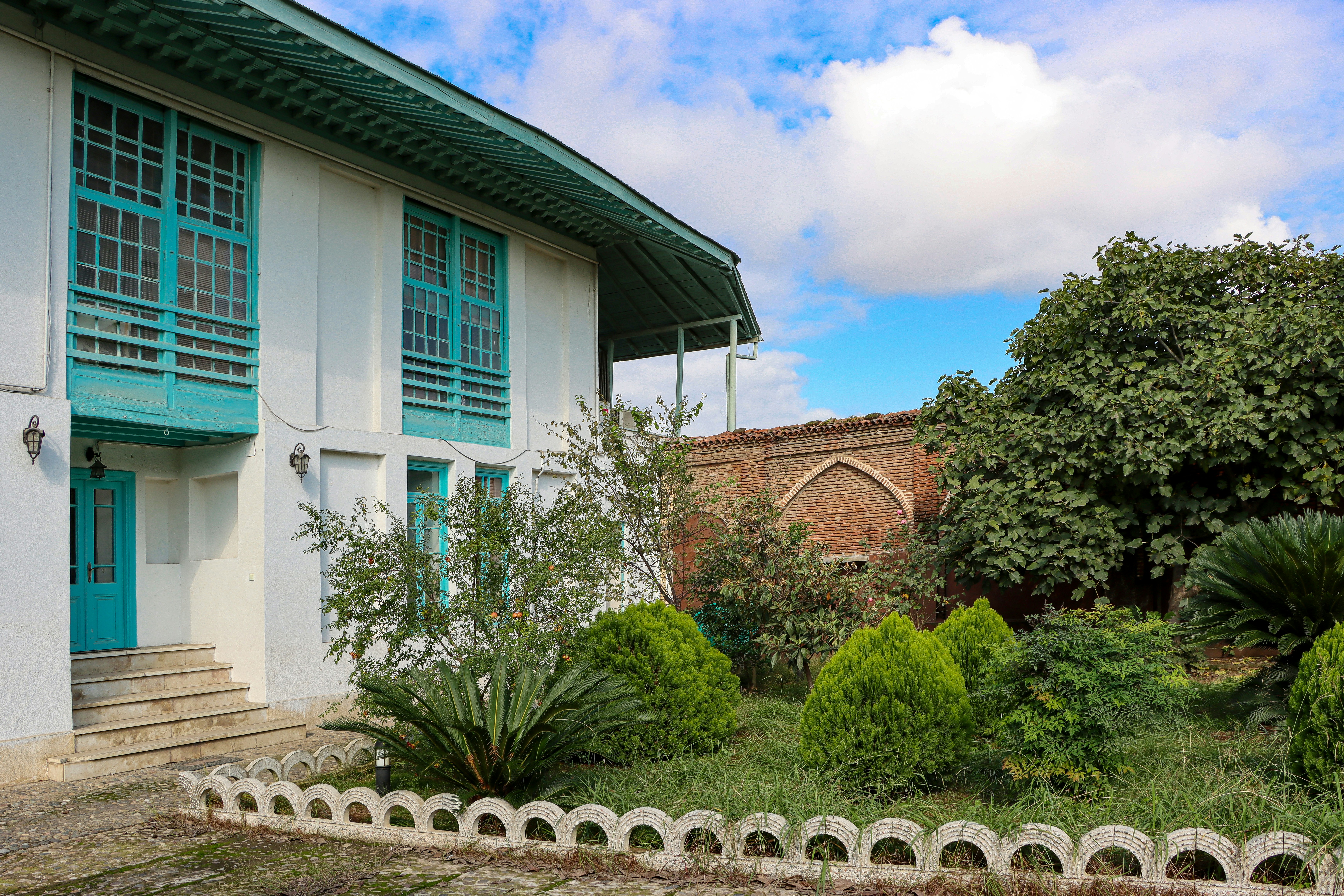 Traditional house in Rasht, Iran. Gilan Cultural House, a historic building in the old urban fabric of Rasht, with turquoise windows, brick walls, and a green courtyard. | White building with teal accents and lush garden.