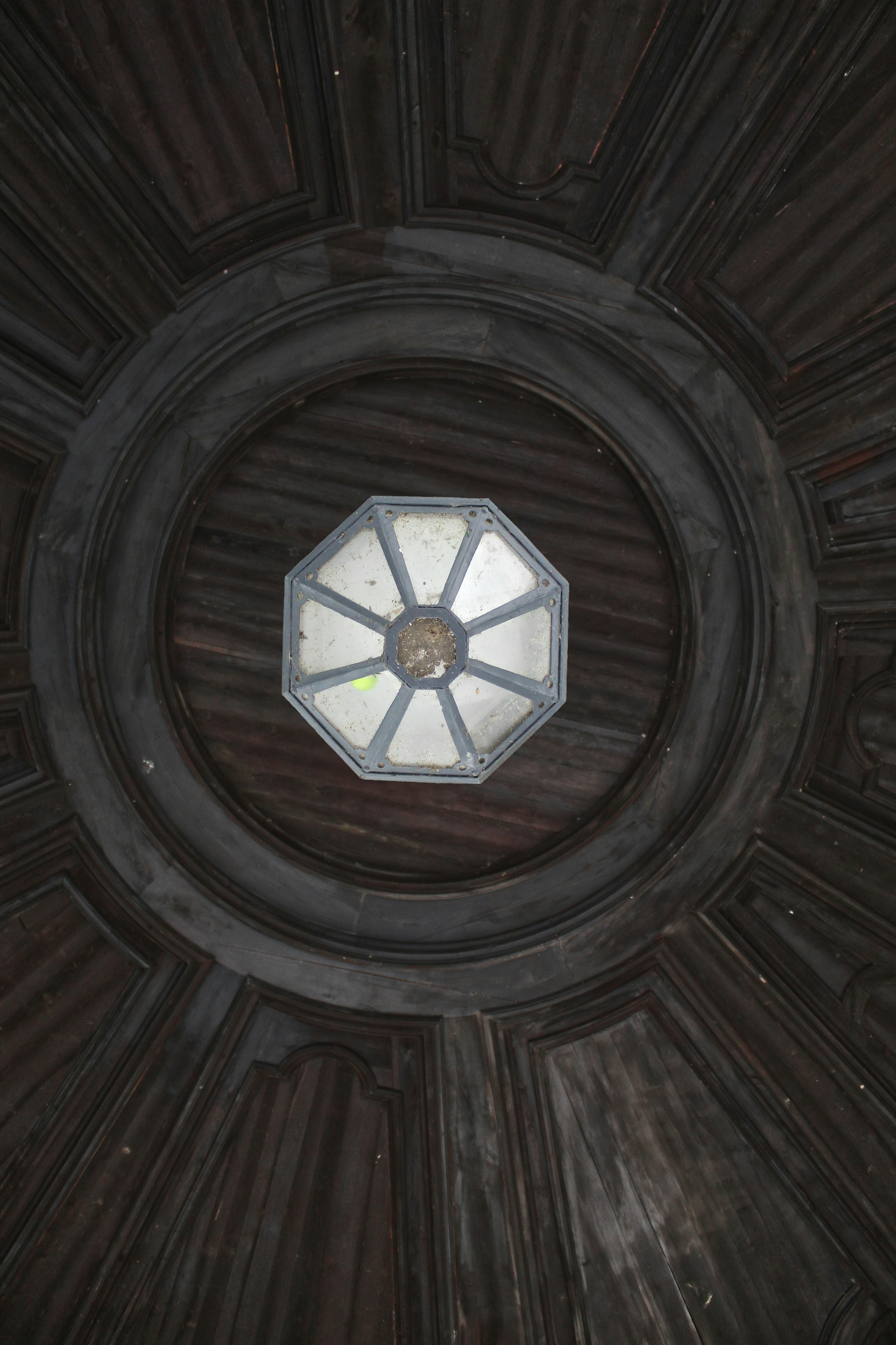 Blick nach oben auf eine alte Kuppel mit einem achteckigen Oberlicht. Das dunkle Holz und die geometrischen Linien erzeugen einen starken Kontrast zum hellen Fenster in der Mitte, das den Raum dezent erhellt. Eine spannende Mischung aus Architektur, Struktur und Zeitspuren. | Octagonal lantern in a dark wooden ceiling