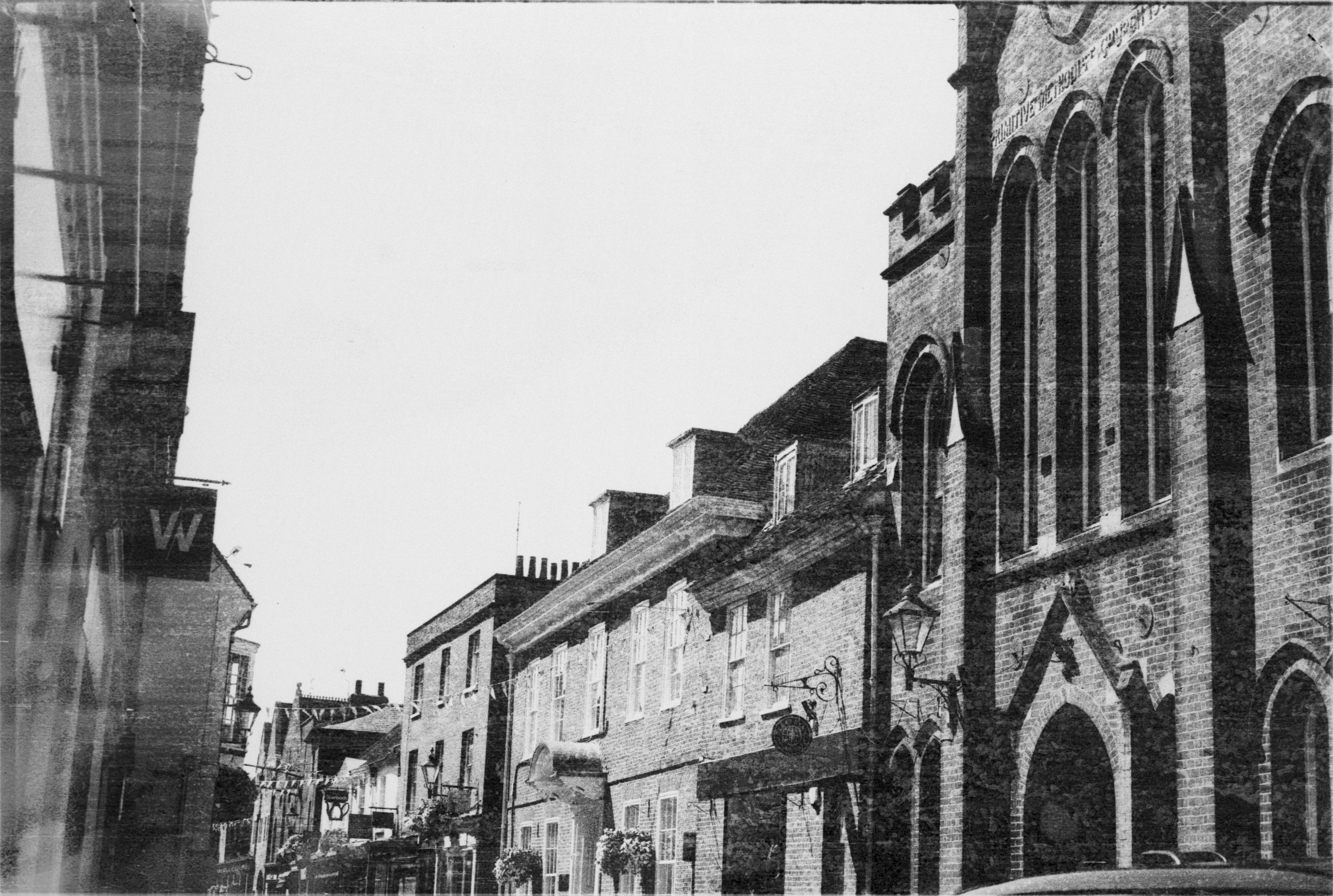 Black and white photograph capturing a historic street lined with brick buildings, showcasing architectural details and a hint of urban life.