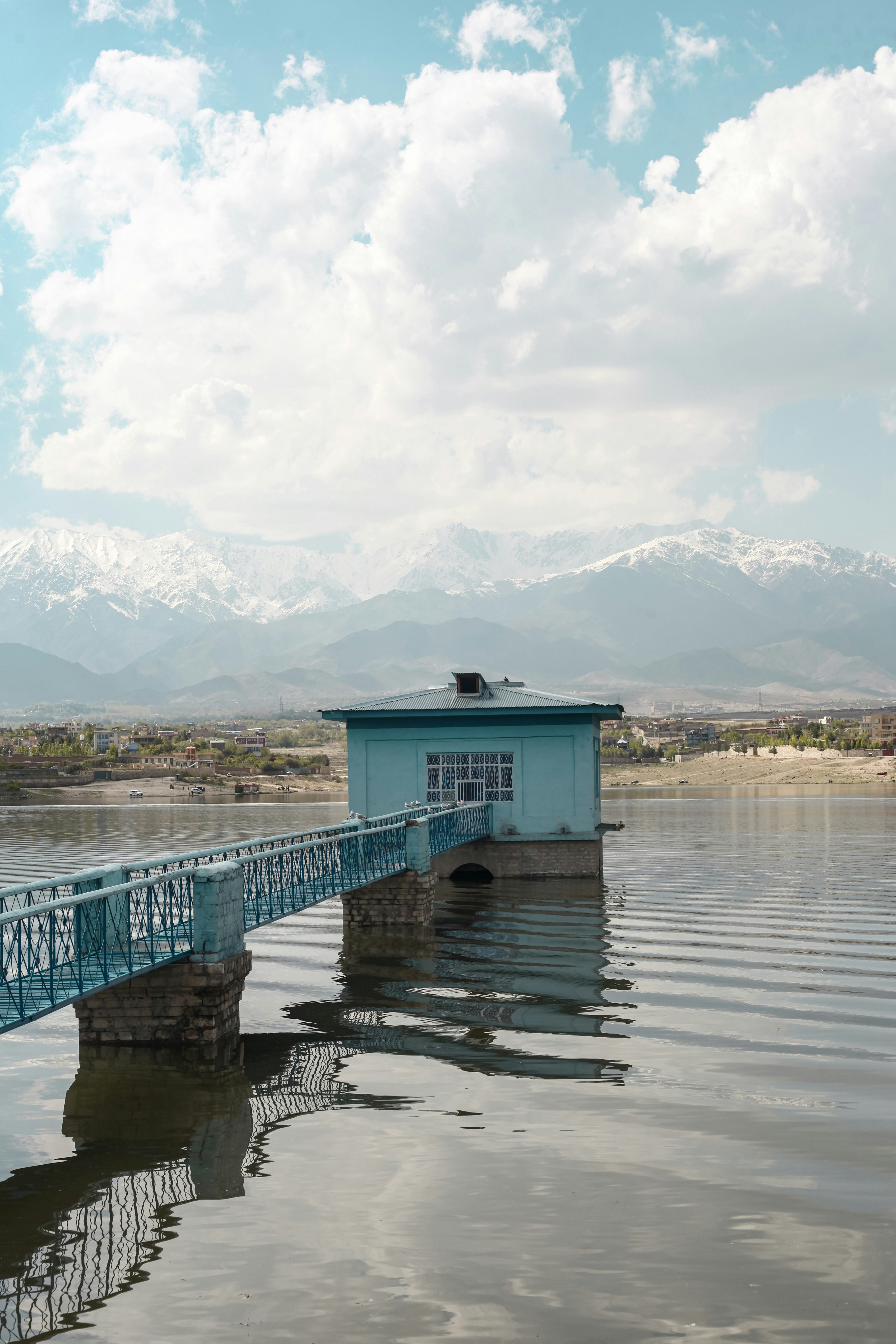 Qargha Lake Park is a beloved recreational destination about 9 kilometers west of Kabul, featuring a man-made reservoir surrounded by gently sloping, verdant hills and mountains, creating a picturesque landscape popular for picnicking, boating, and enjoying local food. Established by the Qargha Dam on the Paghman River, it serves as a tranquil, peaceful escape from the city, particularly popular with families and social gatherings on weekends. | Light blue building on water with snowy mountains