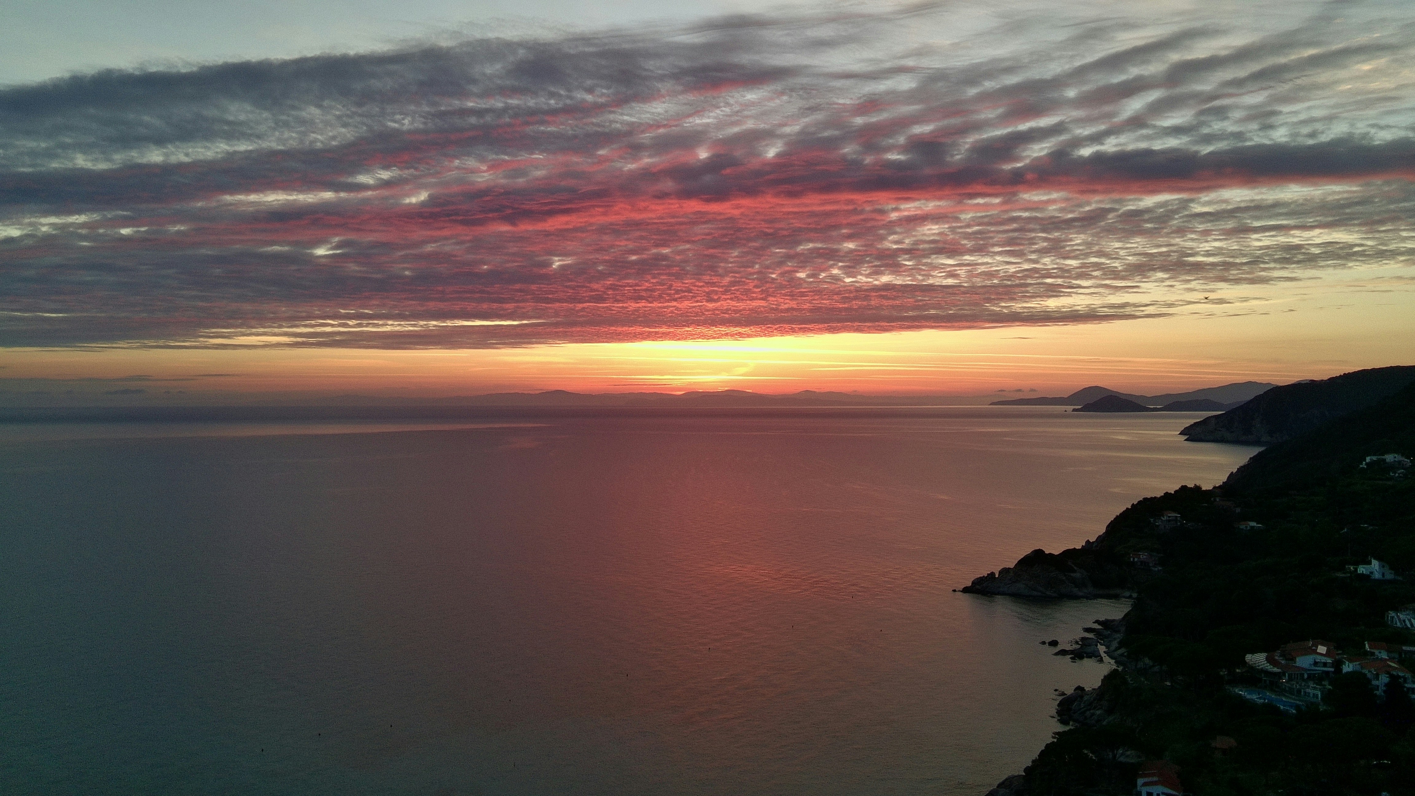 Vibrant sunset reflecting on calm sea waters, framed by distant hills and textured clouds. The scene captures the tranquility of dusk.